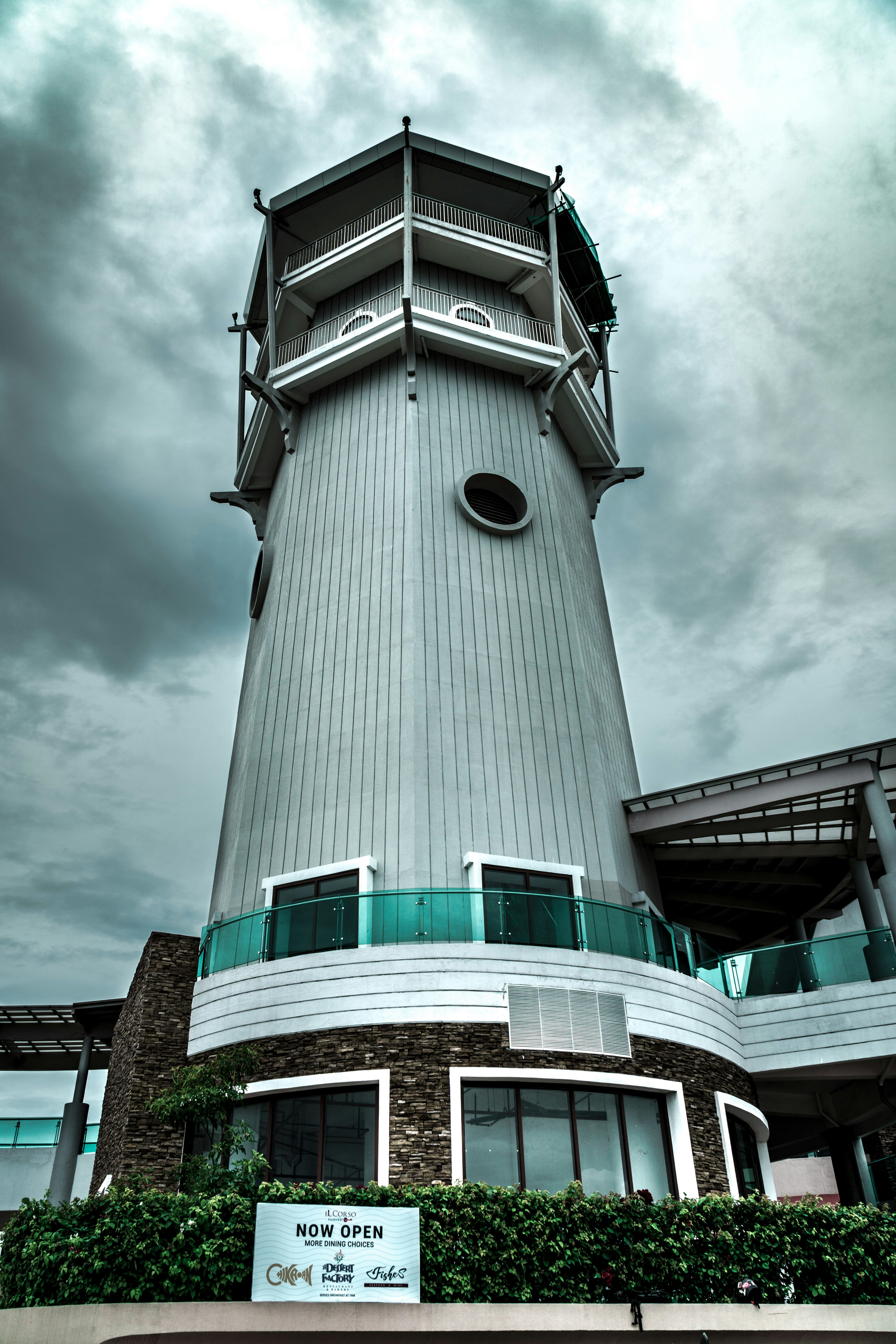 A towering structure with a sleek design, featuring a circular window and a vibrant green terrace, set against a moody sky. The sign at the base indicates a new establishment.