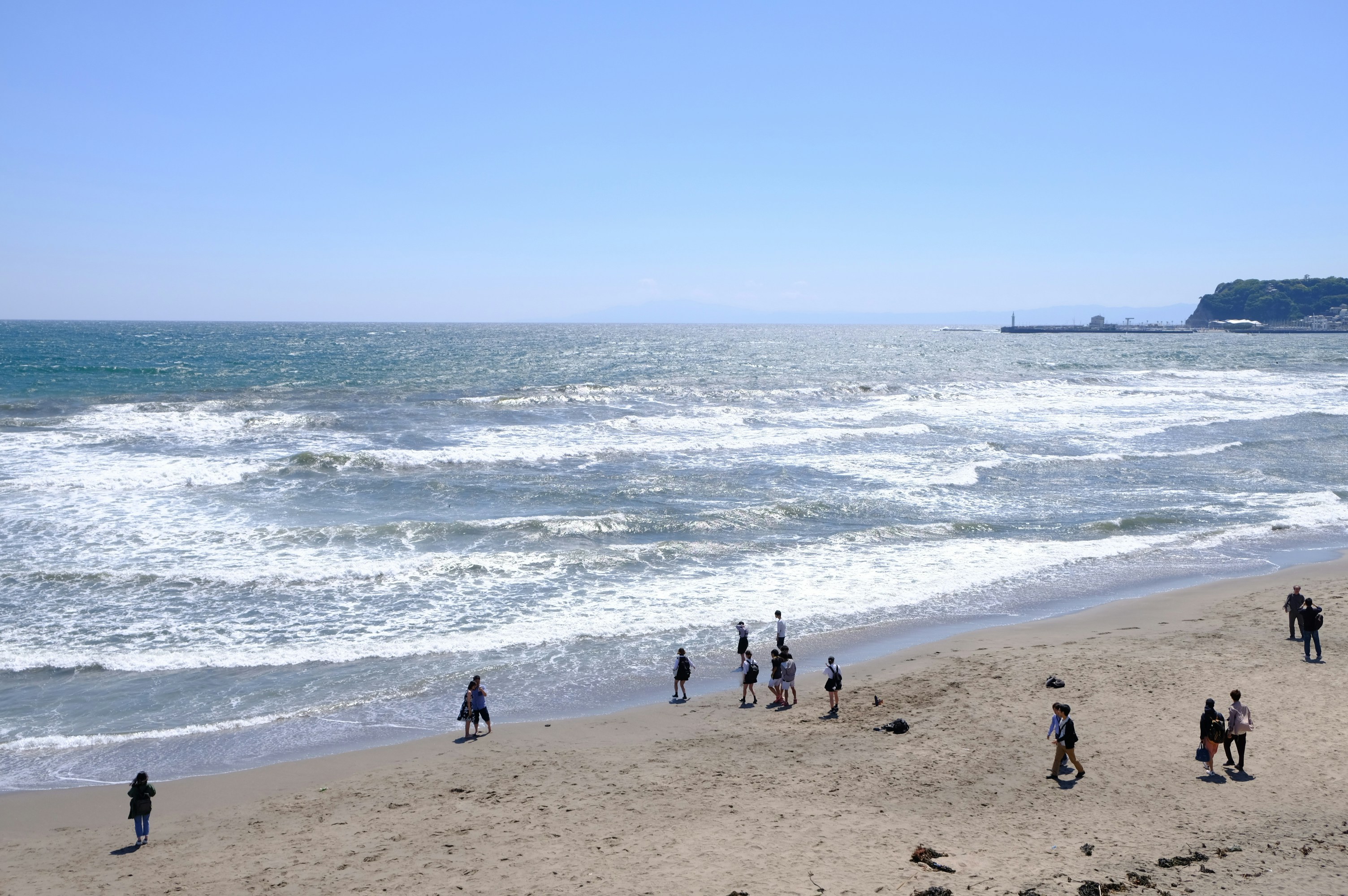 People enjoying a sunny day on a sandy beach with waves crashing ashore and distant cliffs under a clear blue sky.