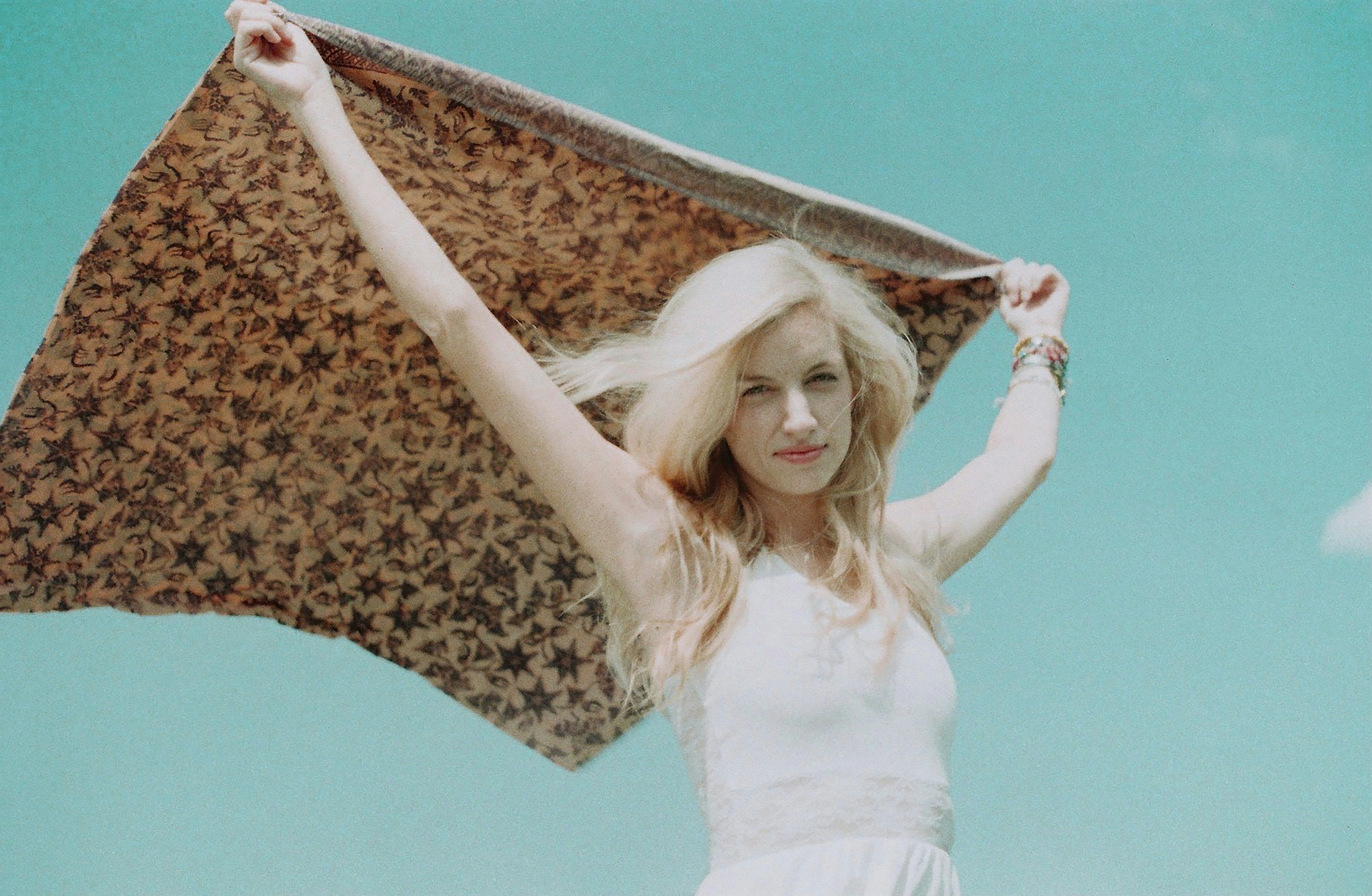 A woman in a flowing white dress holds a patterned scarf aloft against a vibrant blue sky, embodying freedom and joy.