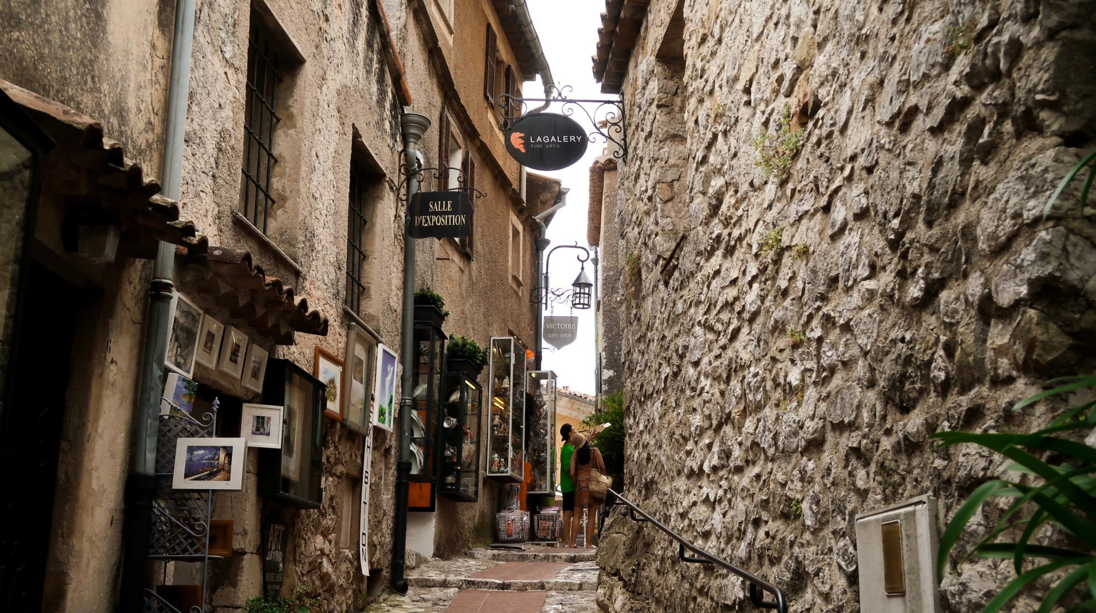 Narrow stone alley with art displays and vintage shop signs, featuring a couple exploring.
