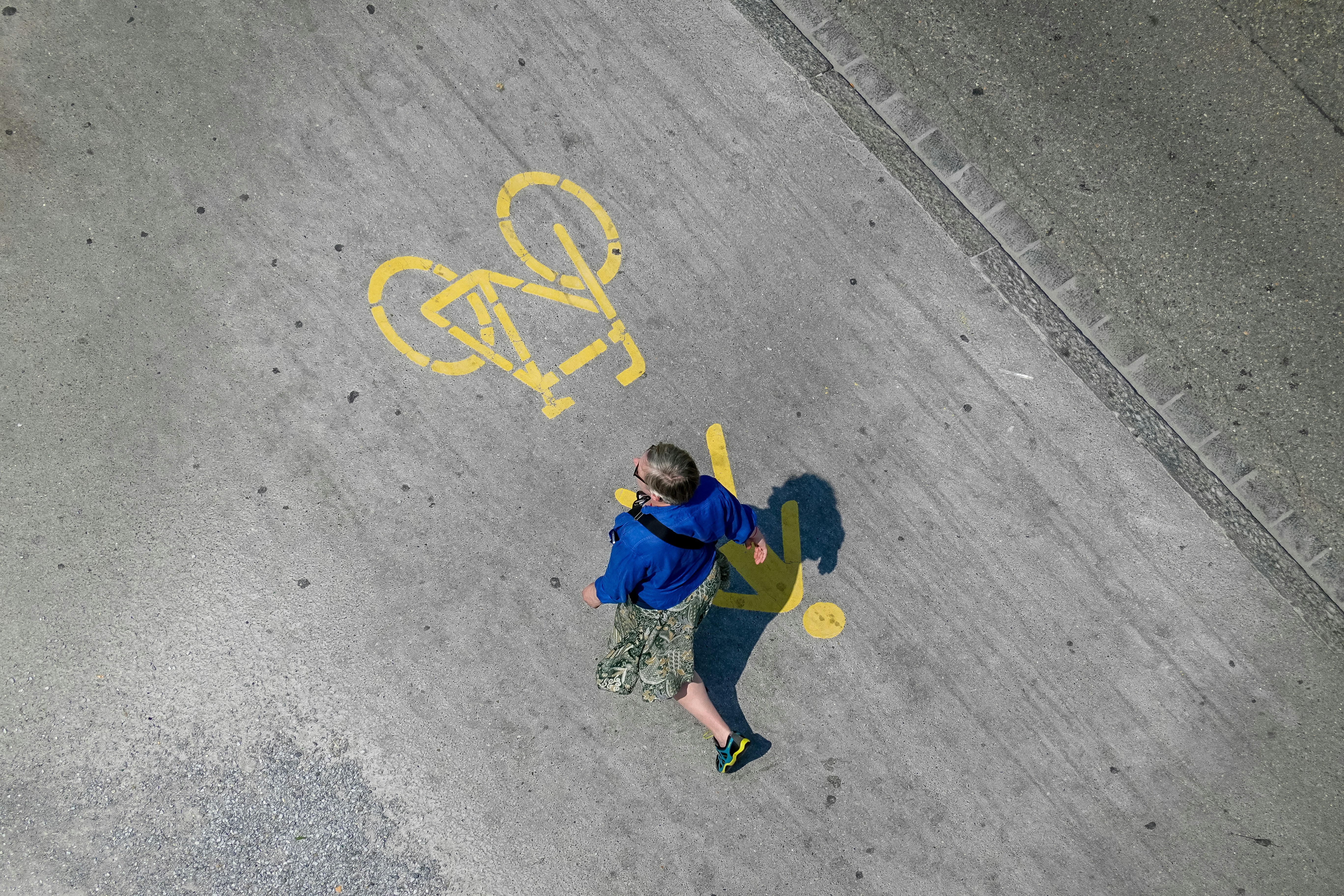 A person in a blue hoodie walks past a yellow bicycle symbol on a concrete surface, casting a shadow on the ground.