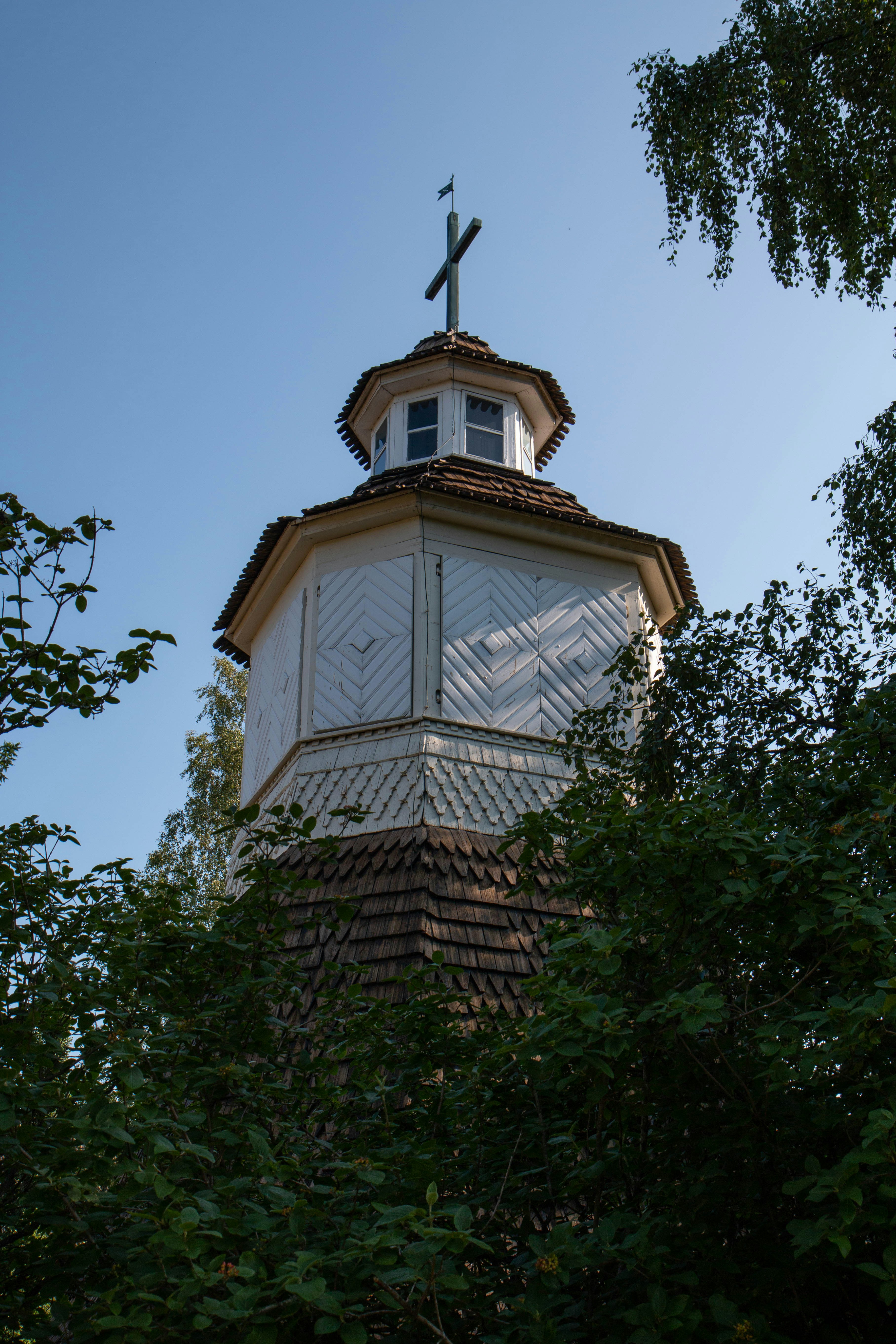 A wooden church tower peeks through lush greenery under a clear blue sky. The intricate design showcases traditional craftsmanship.