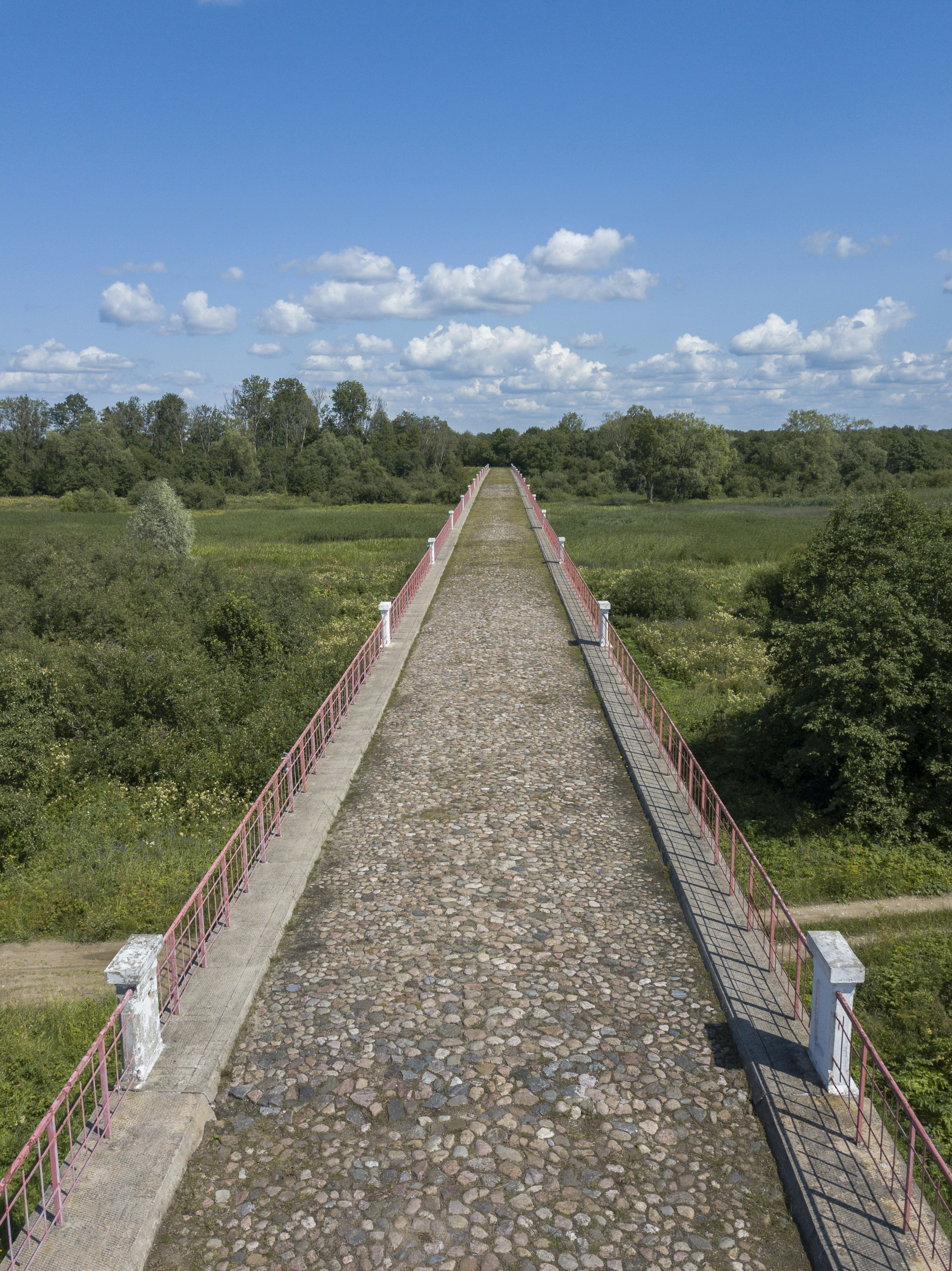 A long, cobblestone bridge stretching into lush greenery under a clear blue sky. The structure invites exploration and contemplation.