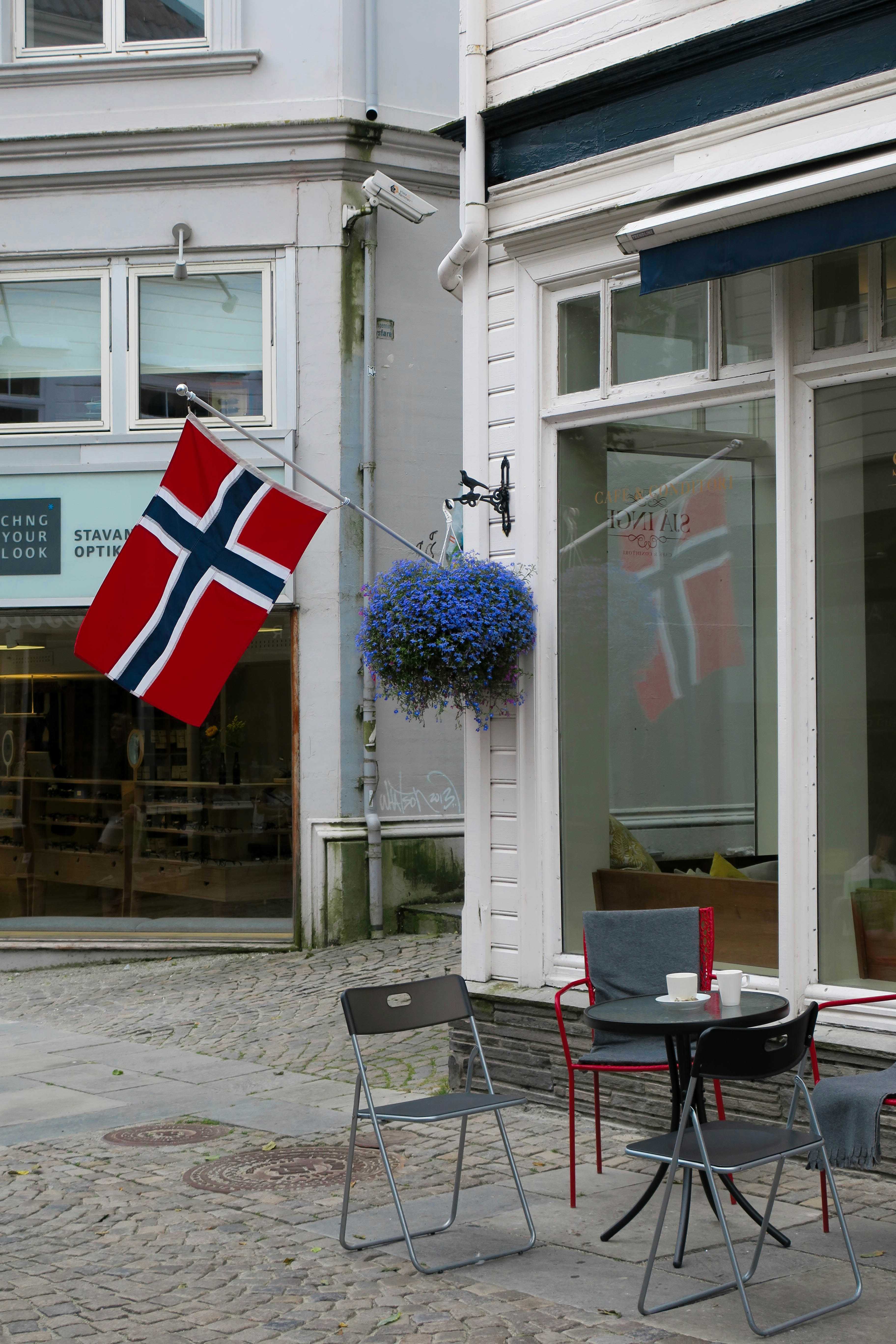 Norwegian flag hangs beside a blue flower basket as a small outdoor café sets up on a cobblestone street. A glass storefront reflects the flag and the simple seating awaits patrons.