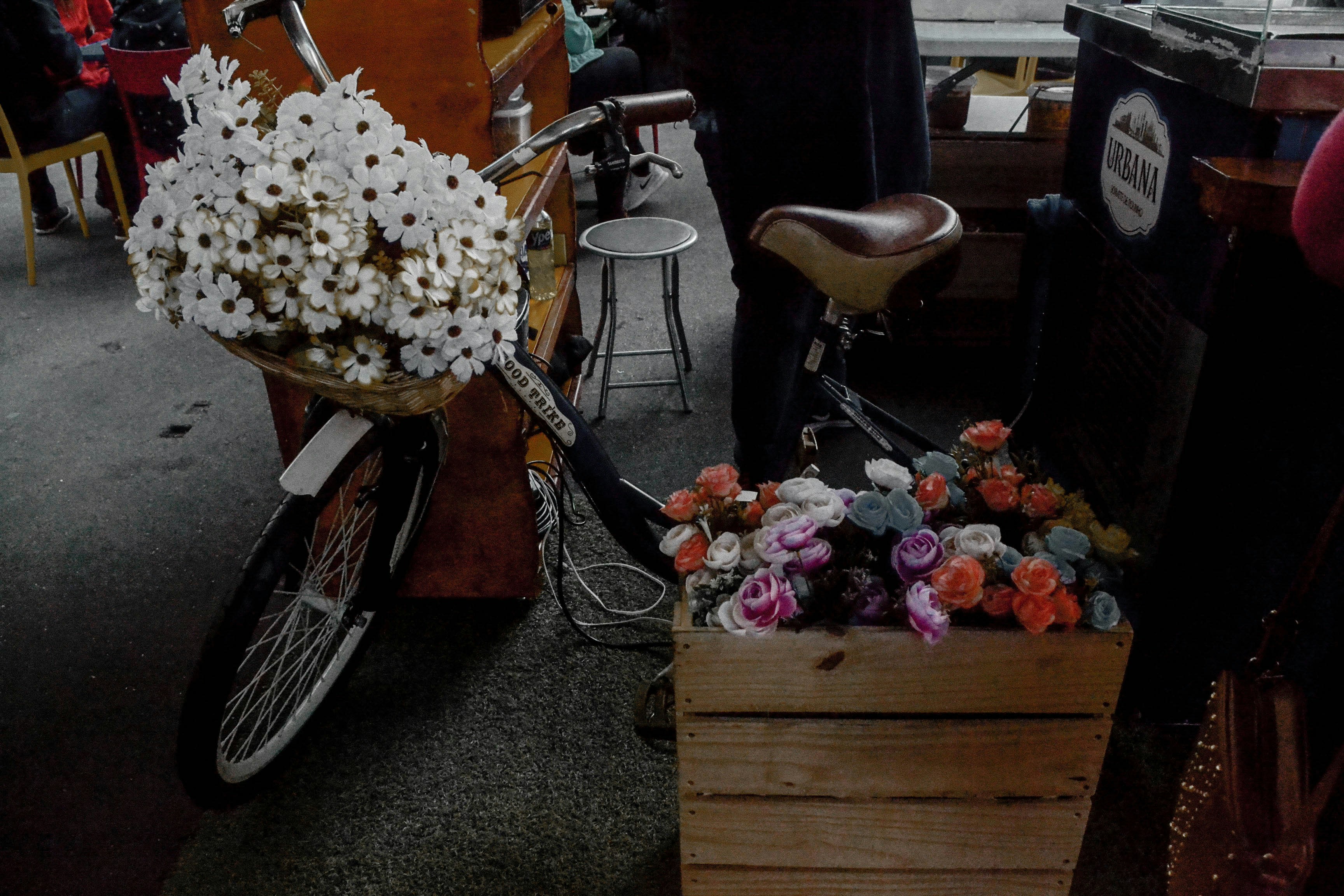 A vintage bicycle adorned with a basket overflowing with white flowers, complemented by a wooden crate filled with colorful roses, set against a bustling market backdrop.