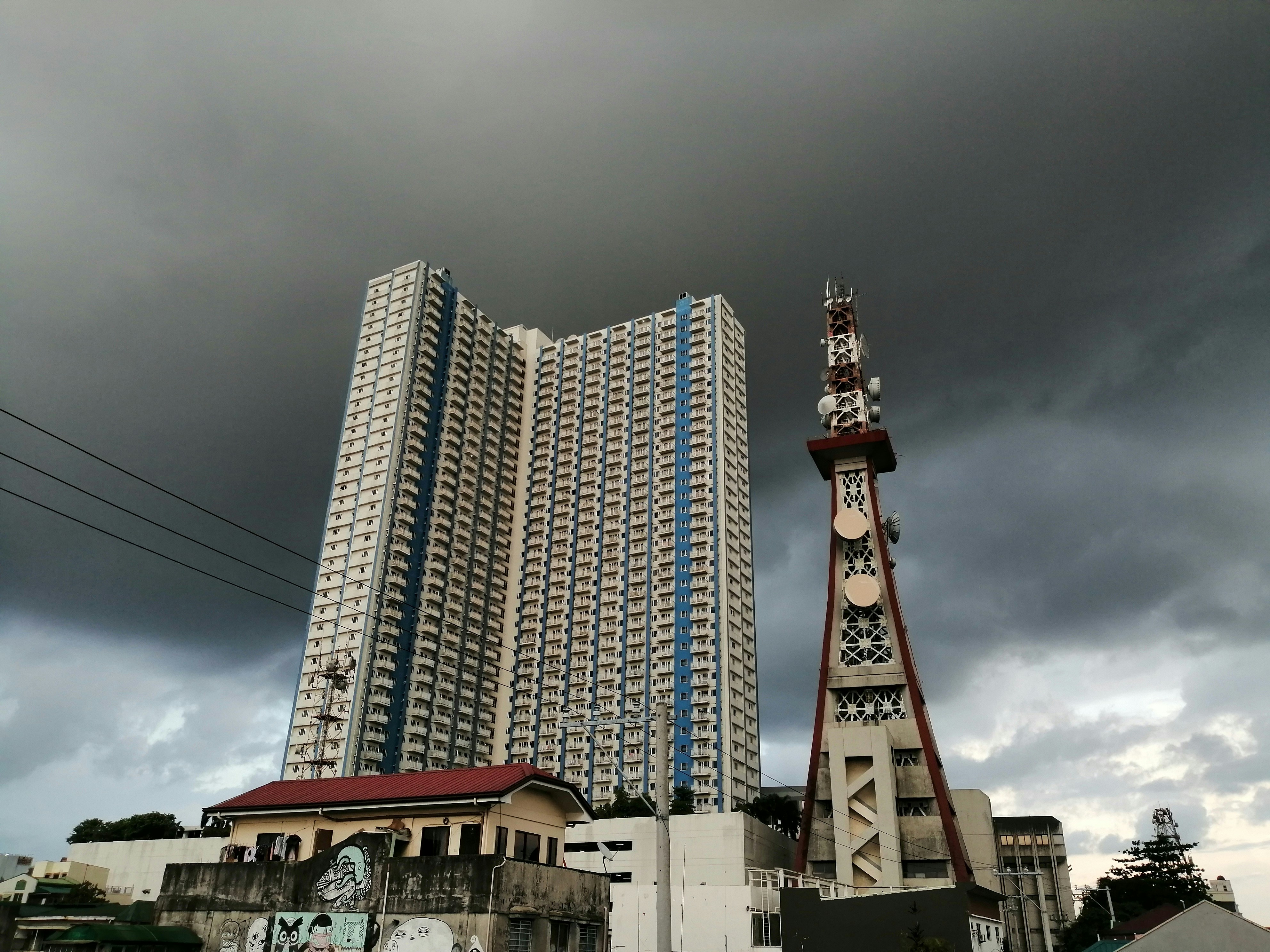 Tall urban skyline with a glass-and-concrete high-rise and a red-and-white communications tower rises beside a low building under a looming storm cloud.