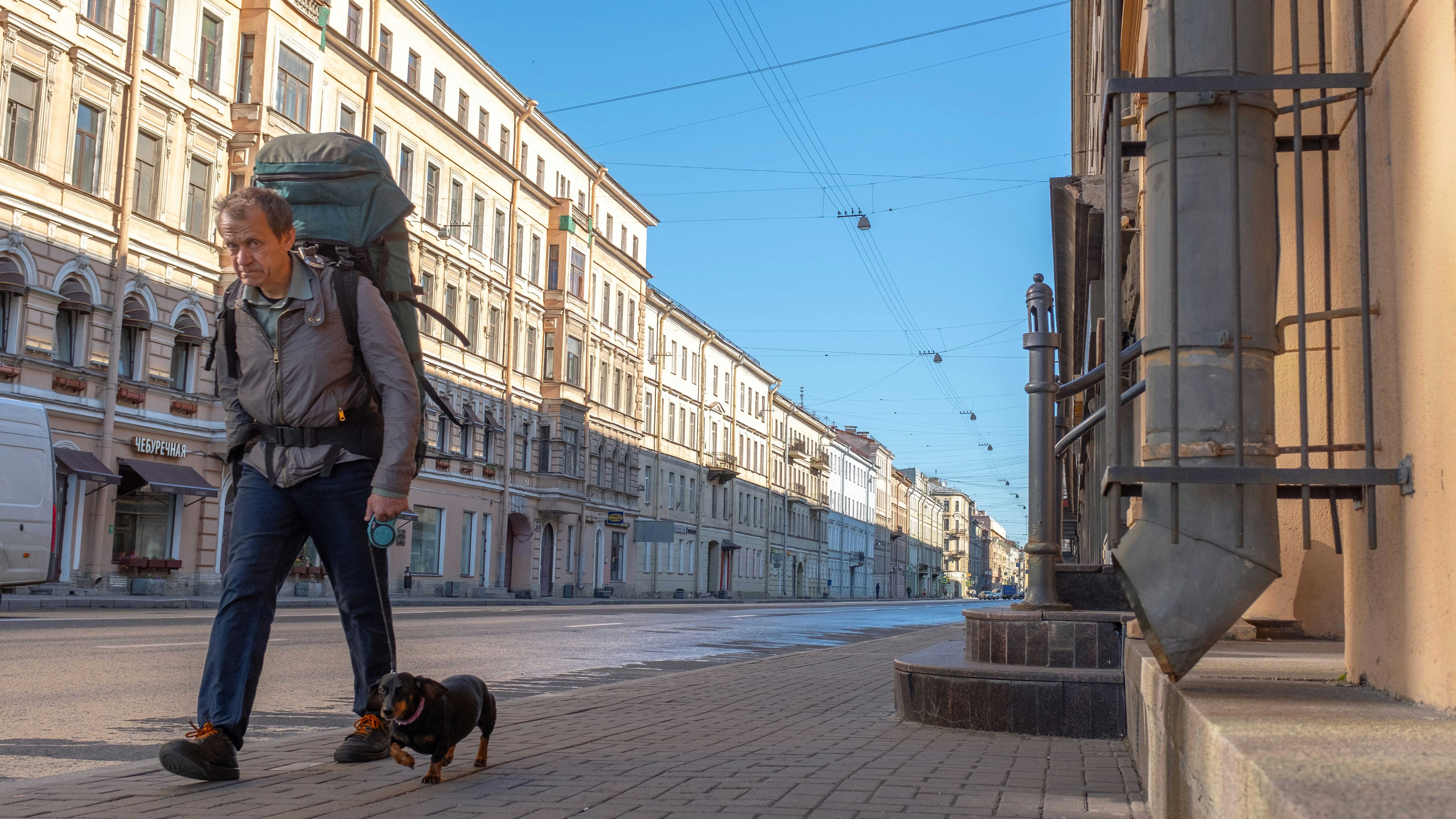 Traveler with a large backpack walks alongside a small dog on a quiet city street, framed by historic architecture under a clear blue sky.