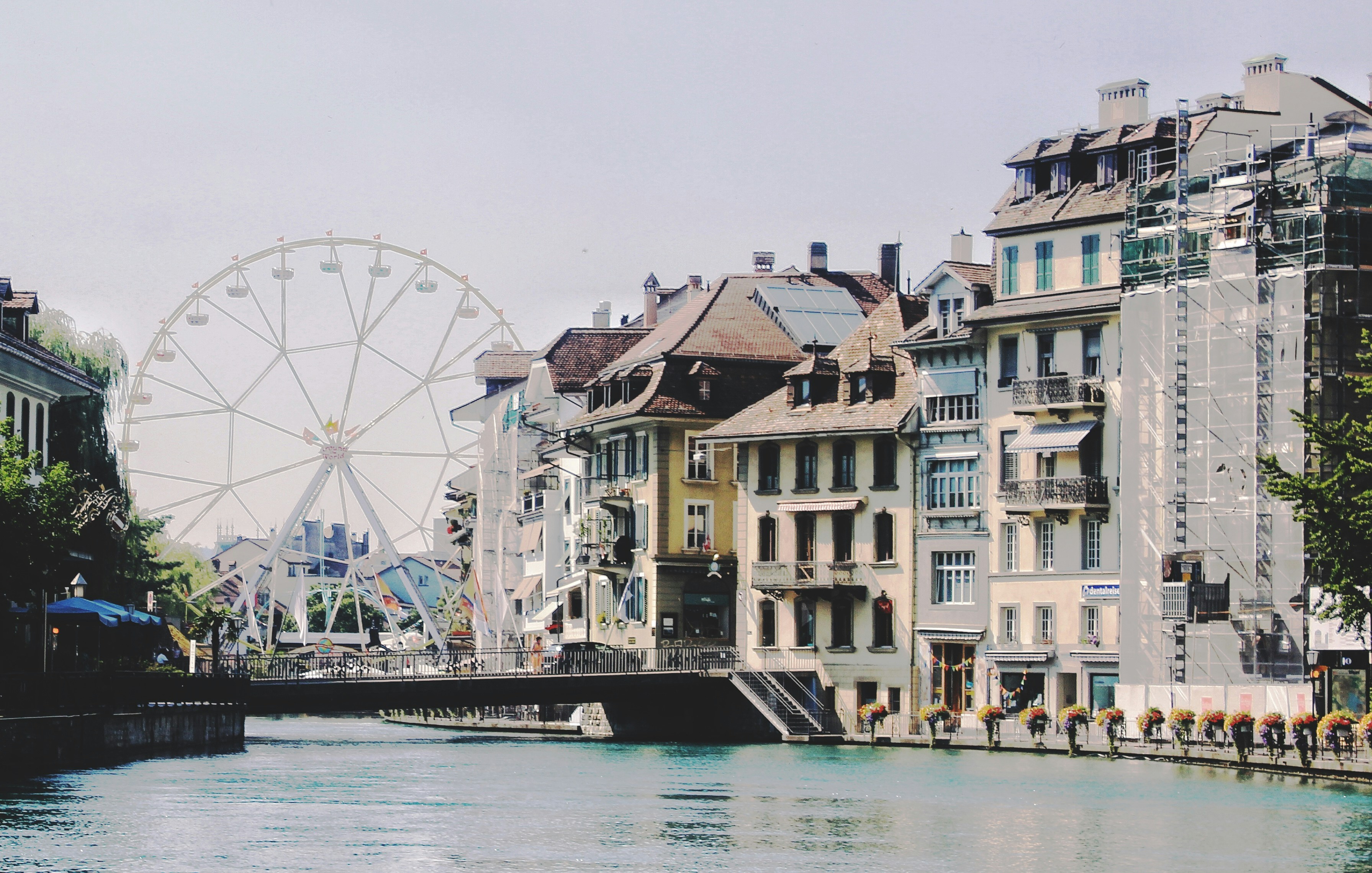 Ferris wheel towering over historic buildings by a tranquil river in Thun.