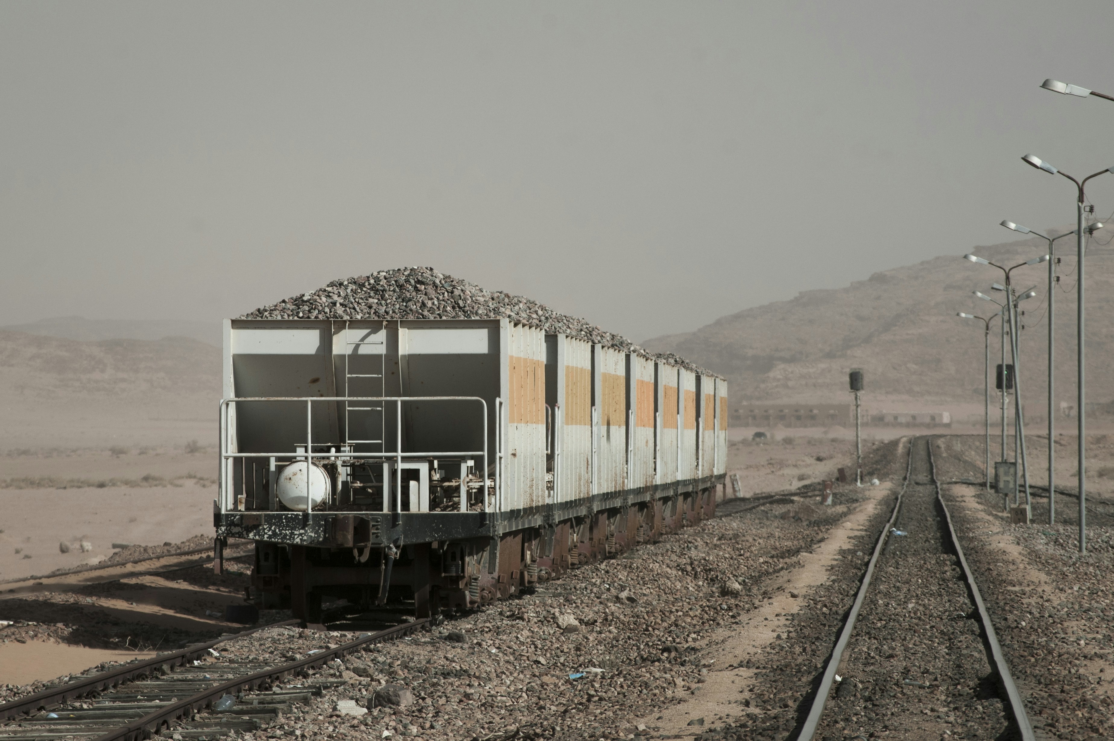 Cargo train loaded with rocks resting on desolate tracks under a hazy sky, evoking a sense of isolation and stillness.