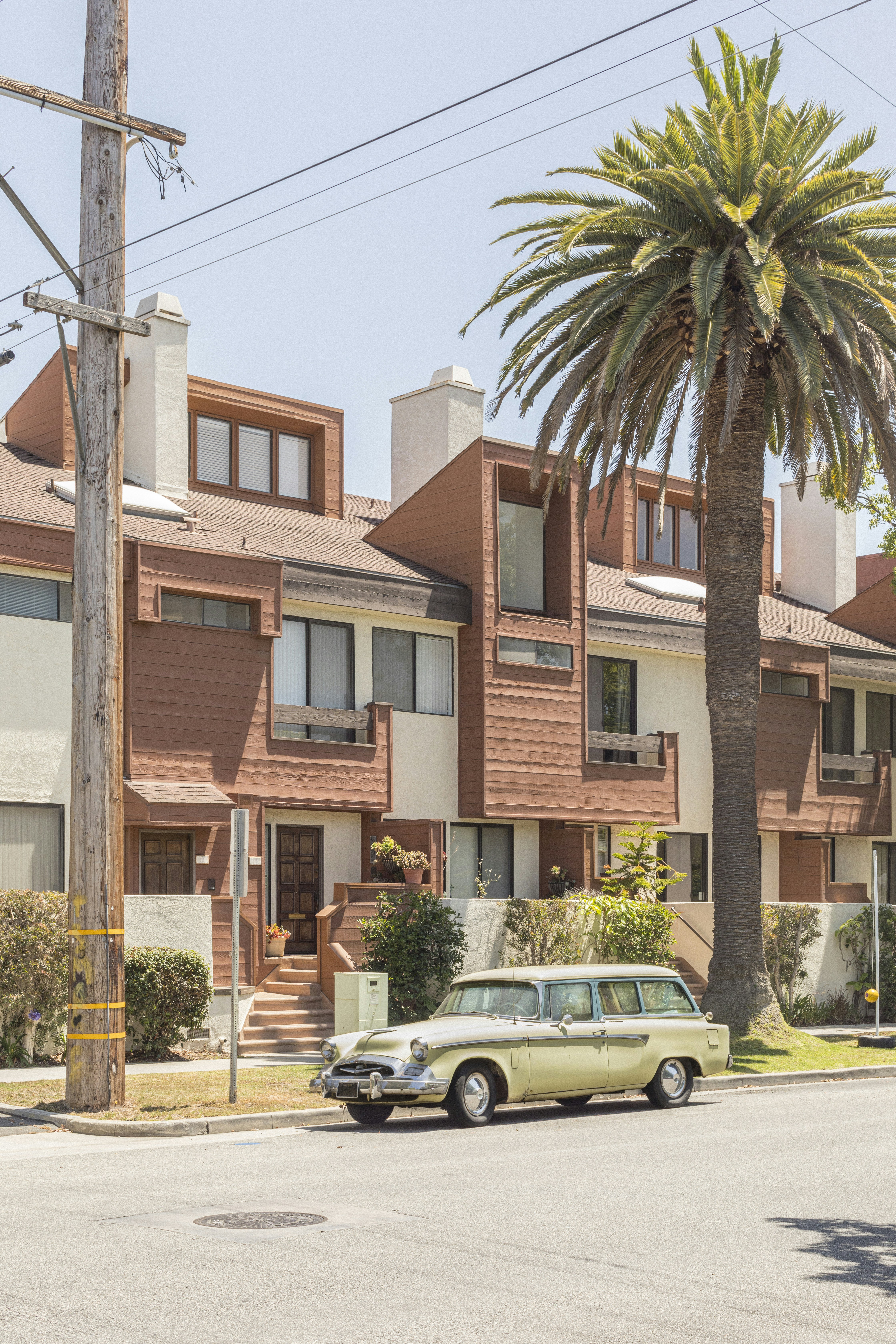 Vintage green car parked in front of townhouses with palm tree and power lines overhead.