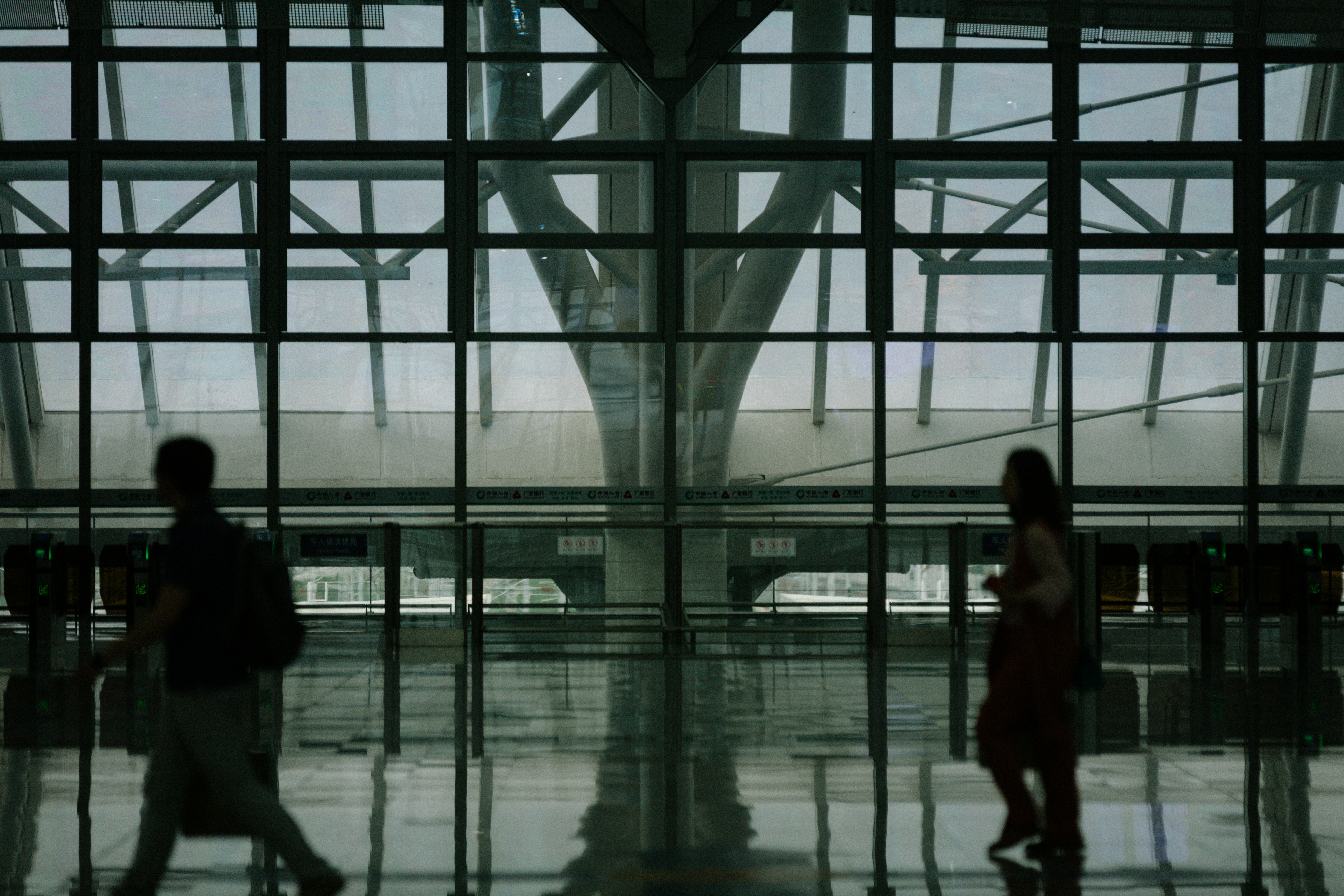 Silhouetted figures of travelers moving through a modern airport terminal, framed by expansive glass structures and reflections on the polished floor.