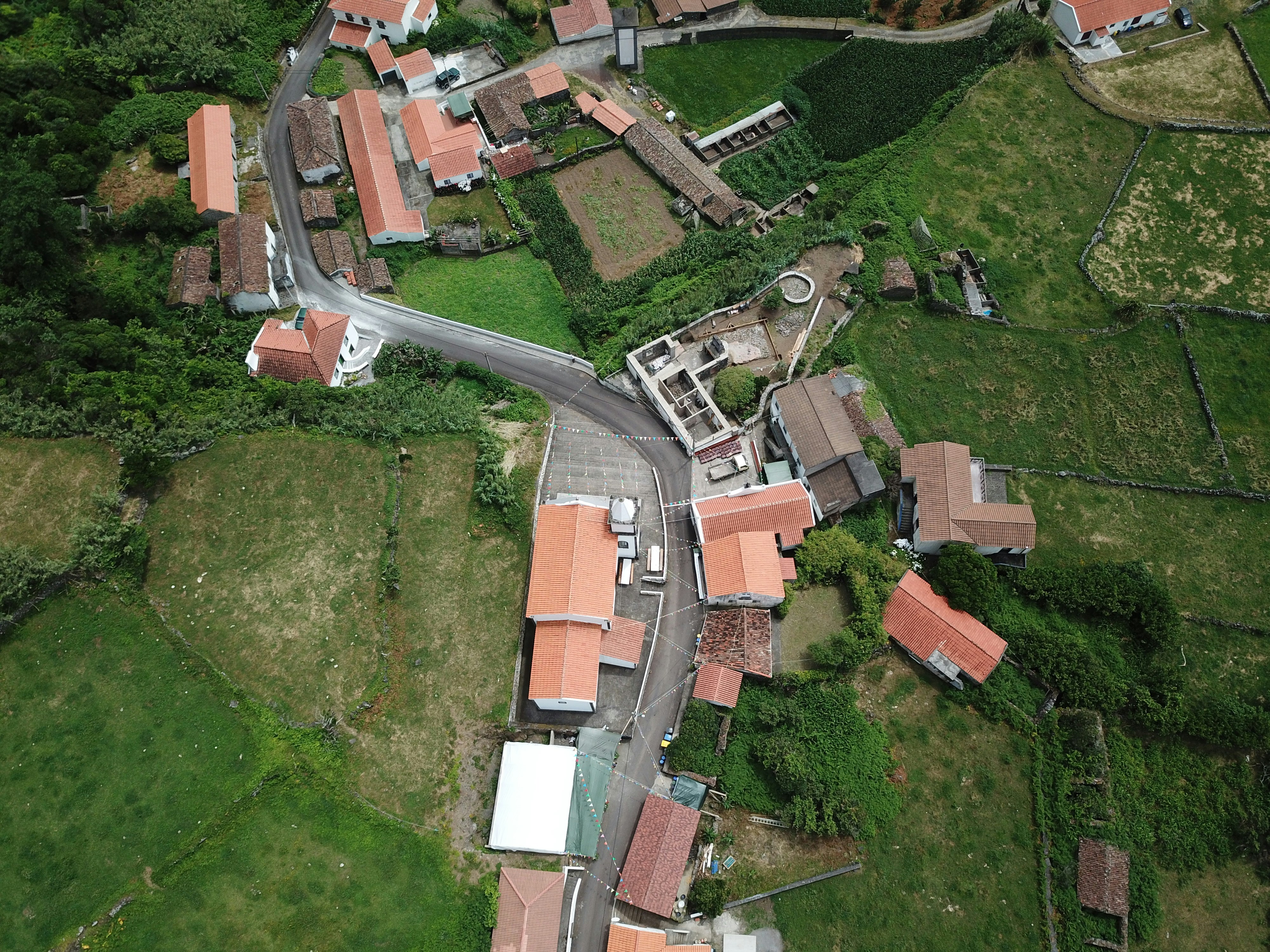 Aerial view of a quaint village with terracotta-roofed houses surrounded by lush green fields and winding roads.
