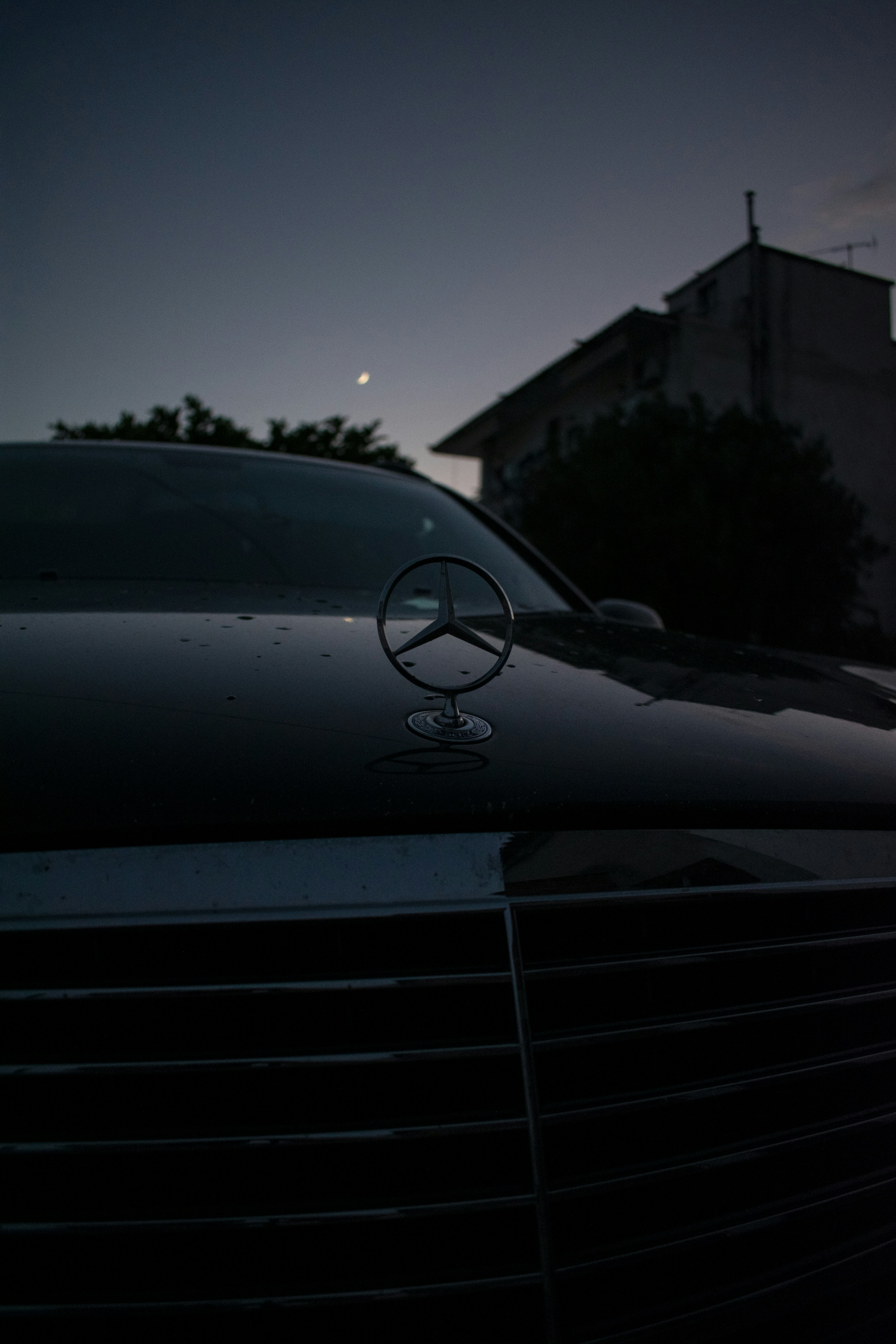 Mercedes-Benz hood ornament glistening softly in the evening light, with a crescent moon visible in the background.