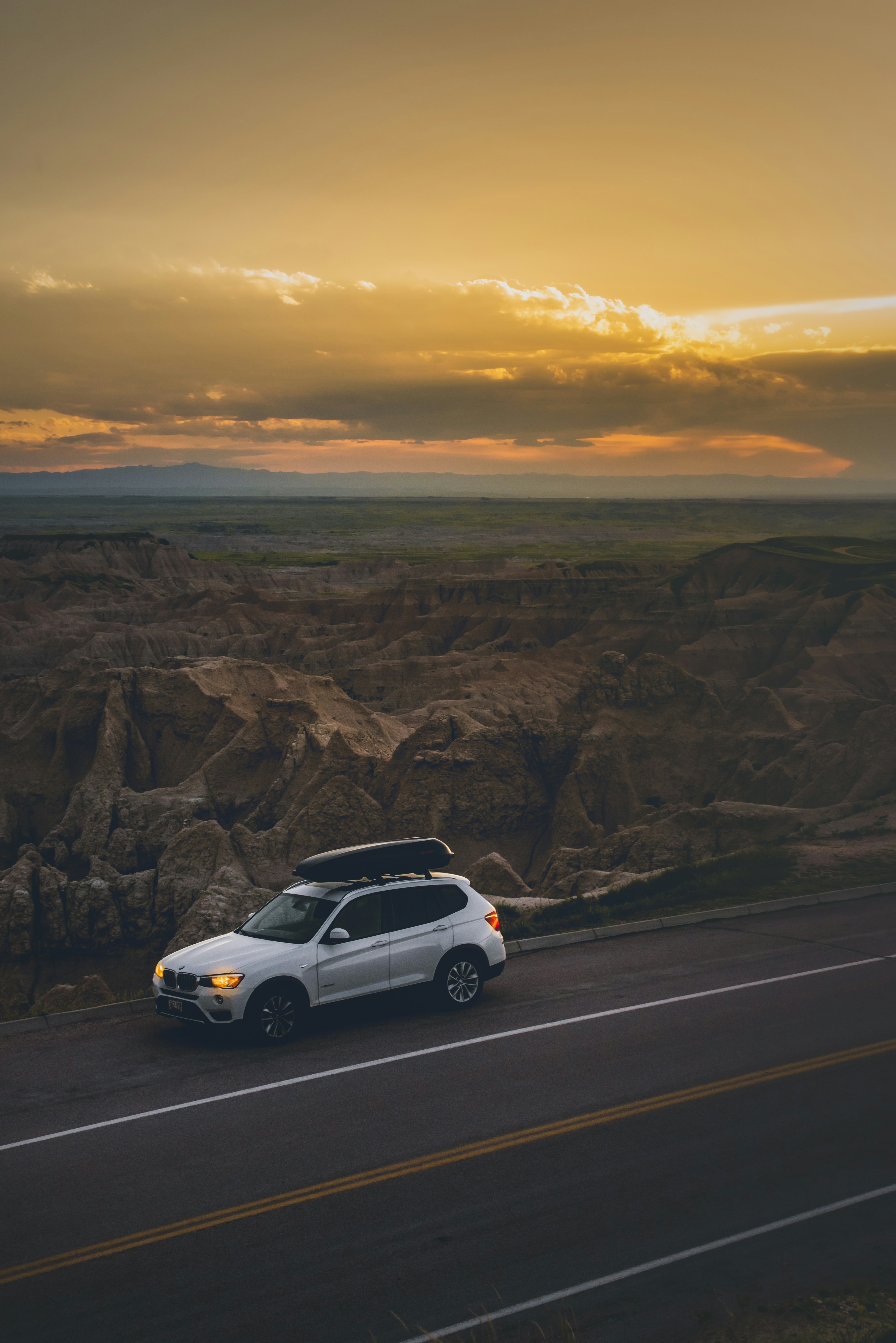 A white SUV parked along a winding road, overlooking the rugged formations of the Badlands at sunset. The warm hues of the sky contrast with the textured landscape.