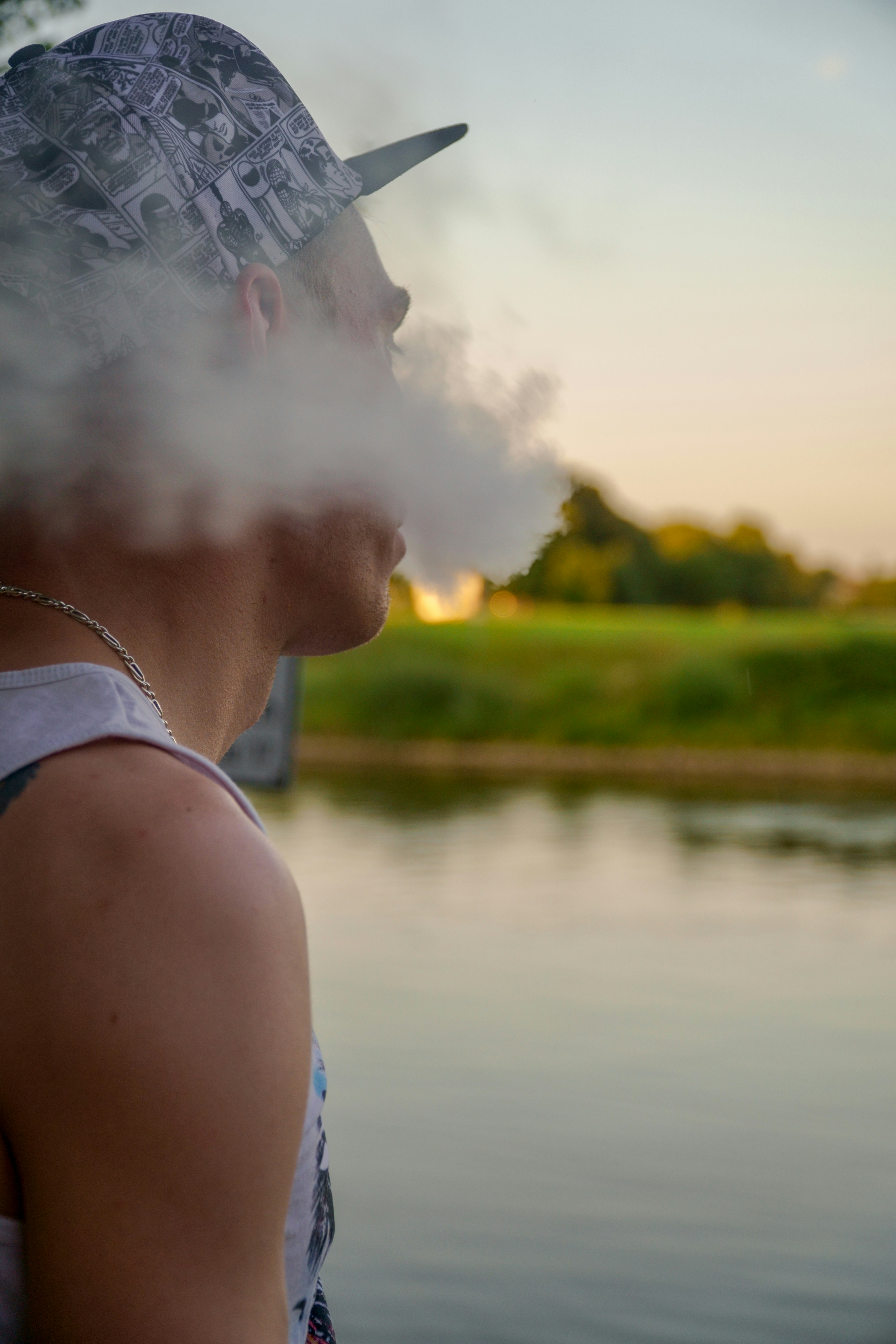 A young man exhales a cloud of vapor while gazing at a serene river landscape during sunset.