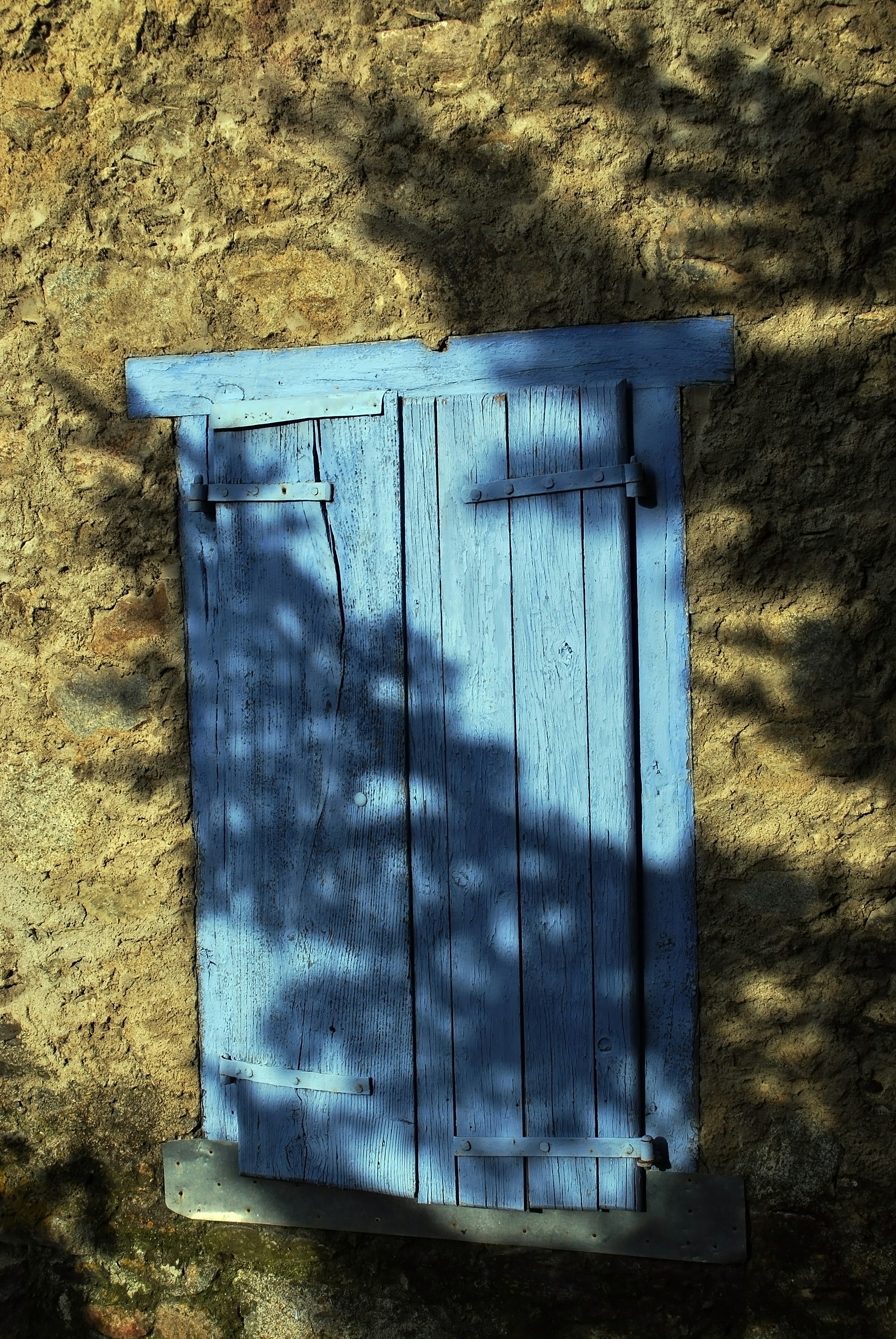 Weathered blue shutters casting shadows on a textured stone wall, evoking a sense of history and tranquility.