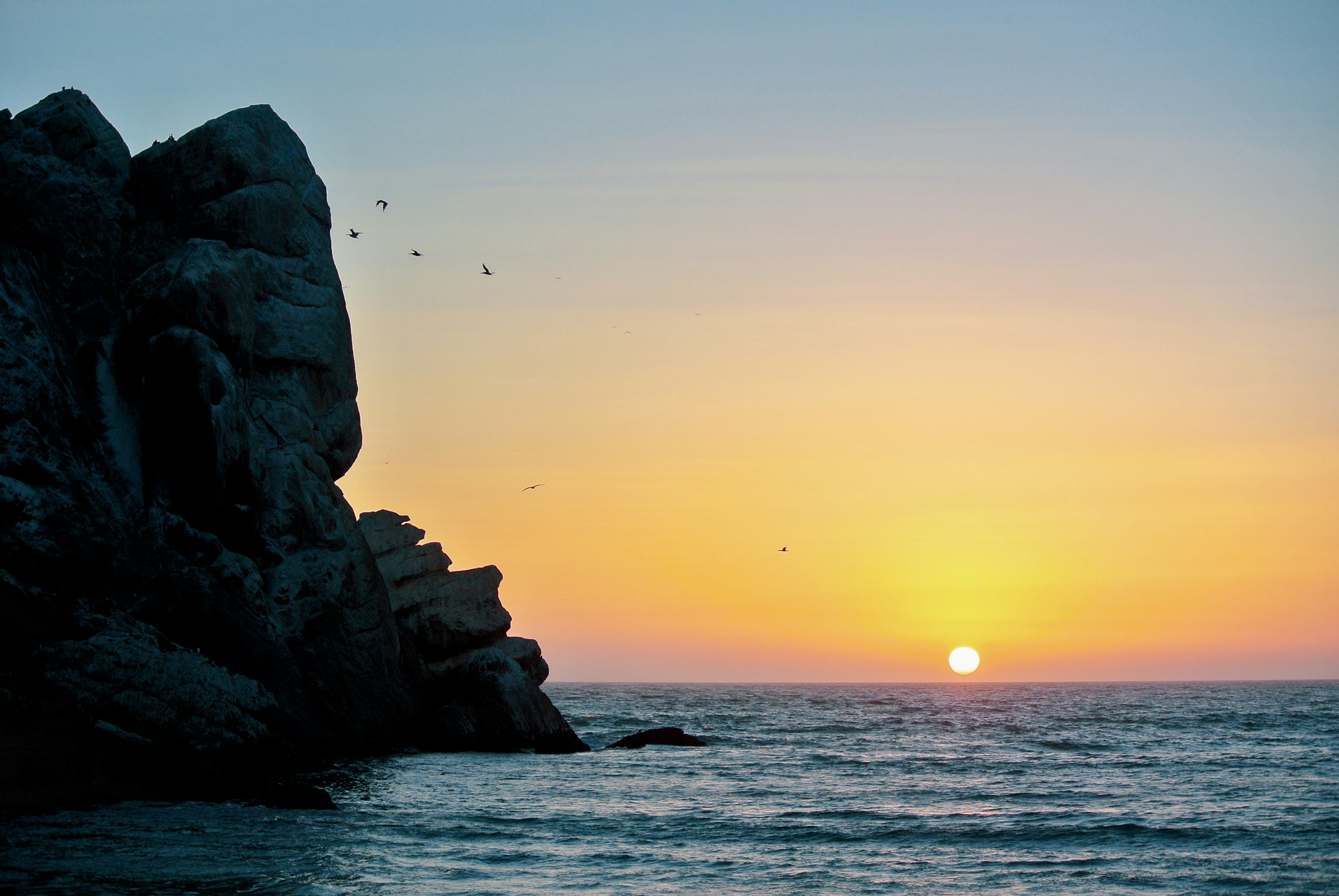 Silhouetted rock formation against a vibrant sunset over the ocean, with birds soaring in the sky. The scene captures the tranquil transition from day to night.