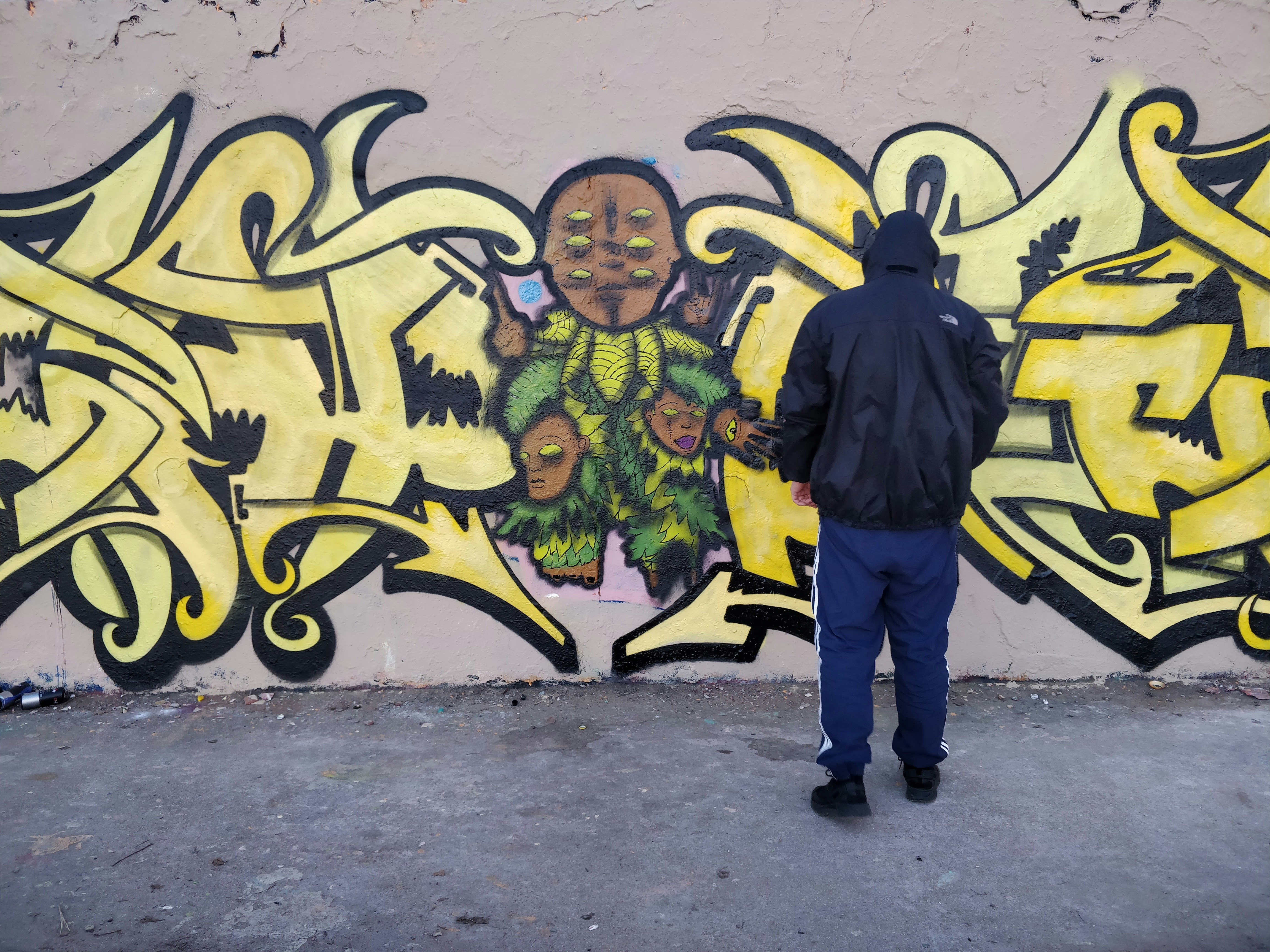 Person in a hooded jacket standing in front of vibrant graffiti featuring abstract yellow designs and a central meditative figure.