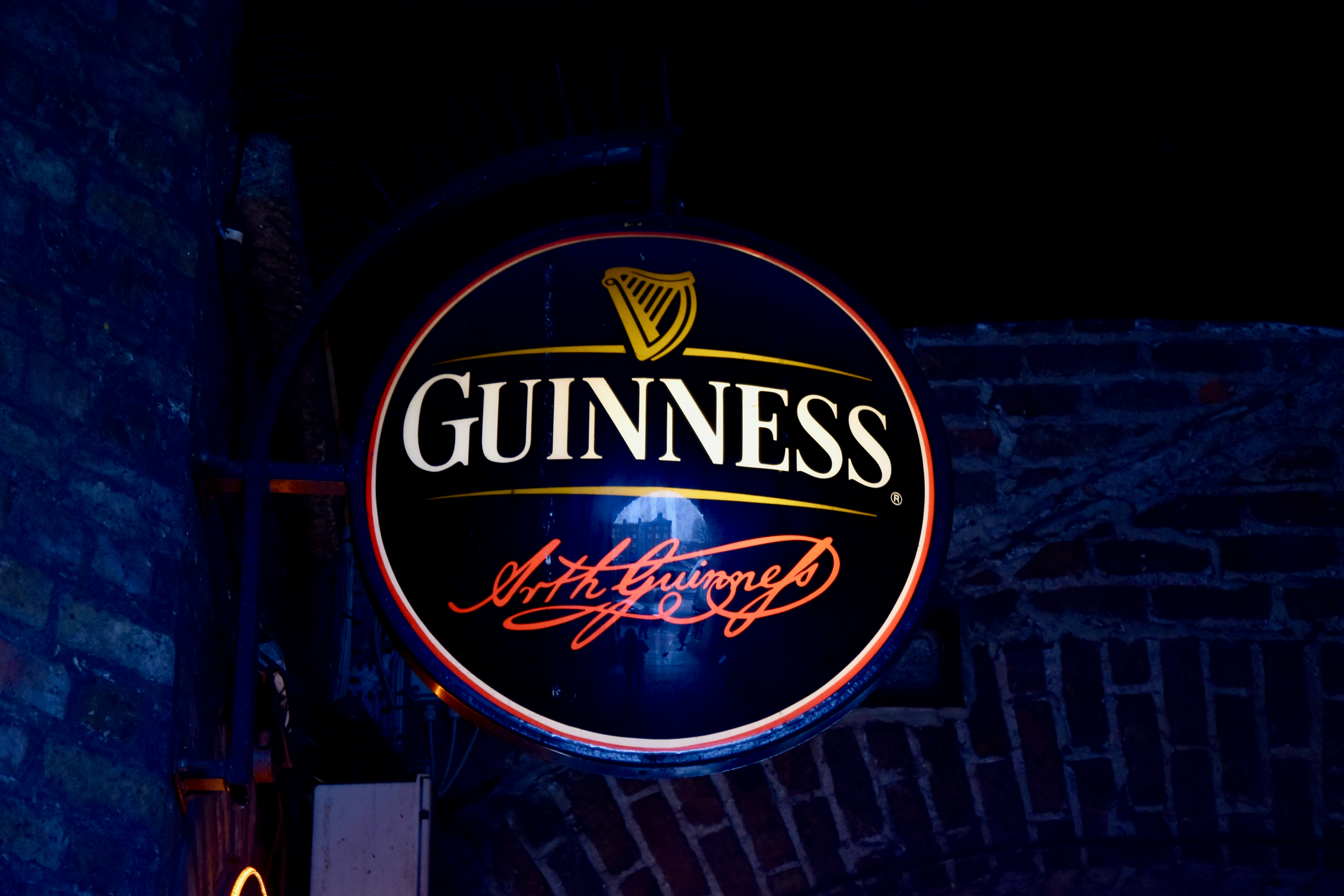 Illuminated Guinness sign against a textured brick background, showcasing the iconic logo and vibrant colors.