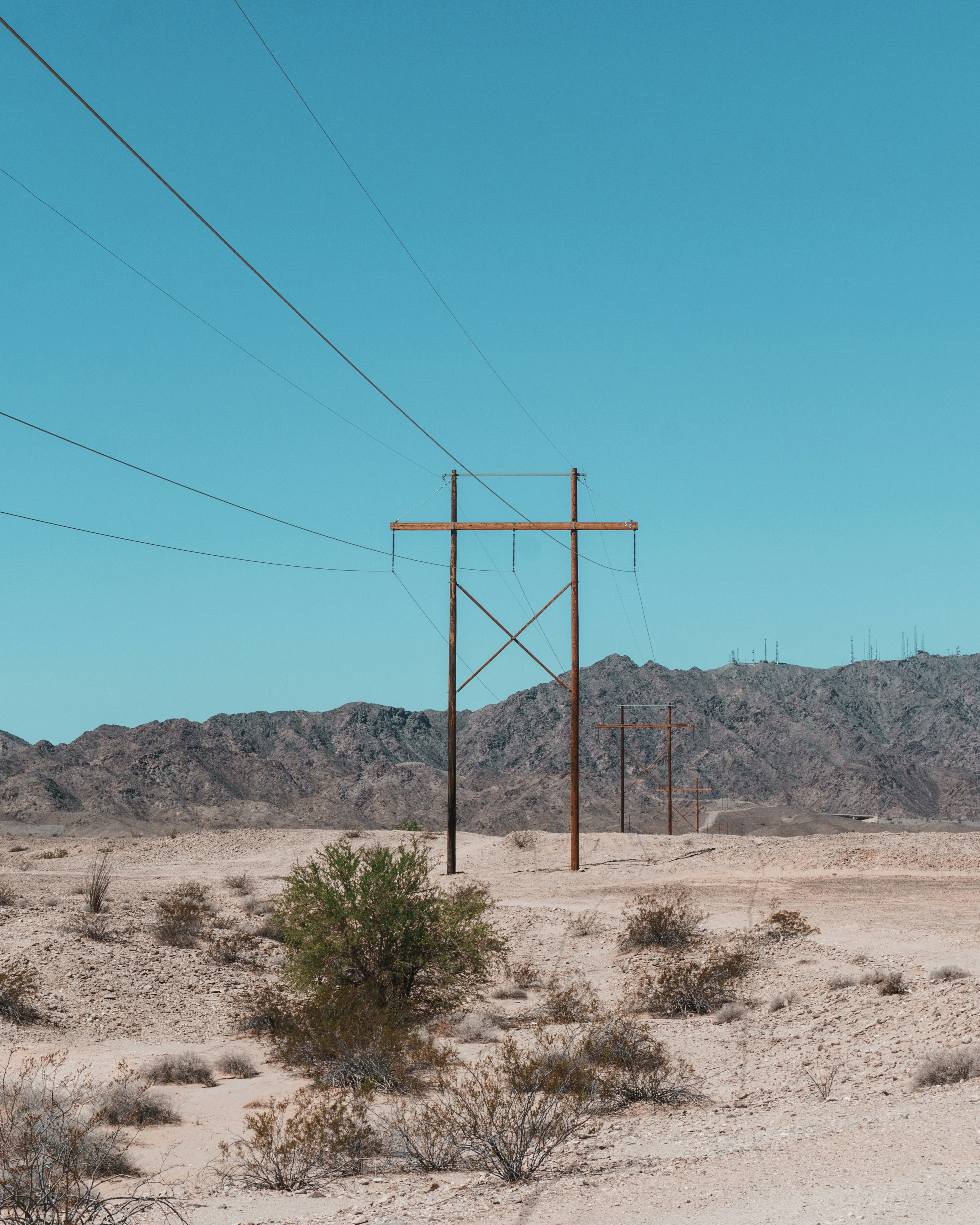 Rusty power lines stretch across a stark desert landscape, framed by distant mountains under a clear blue sky.