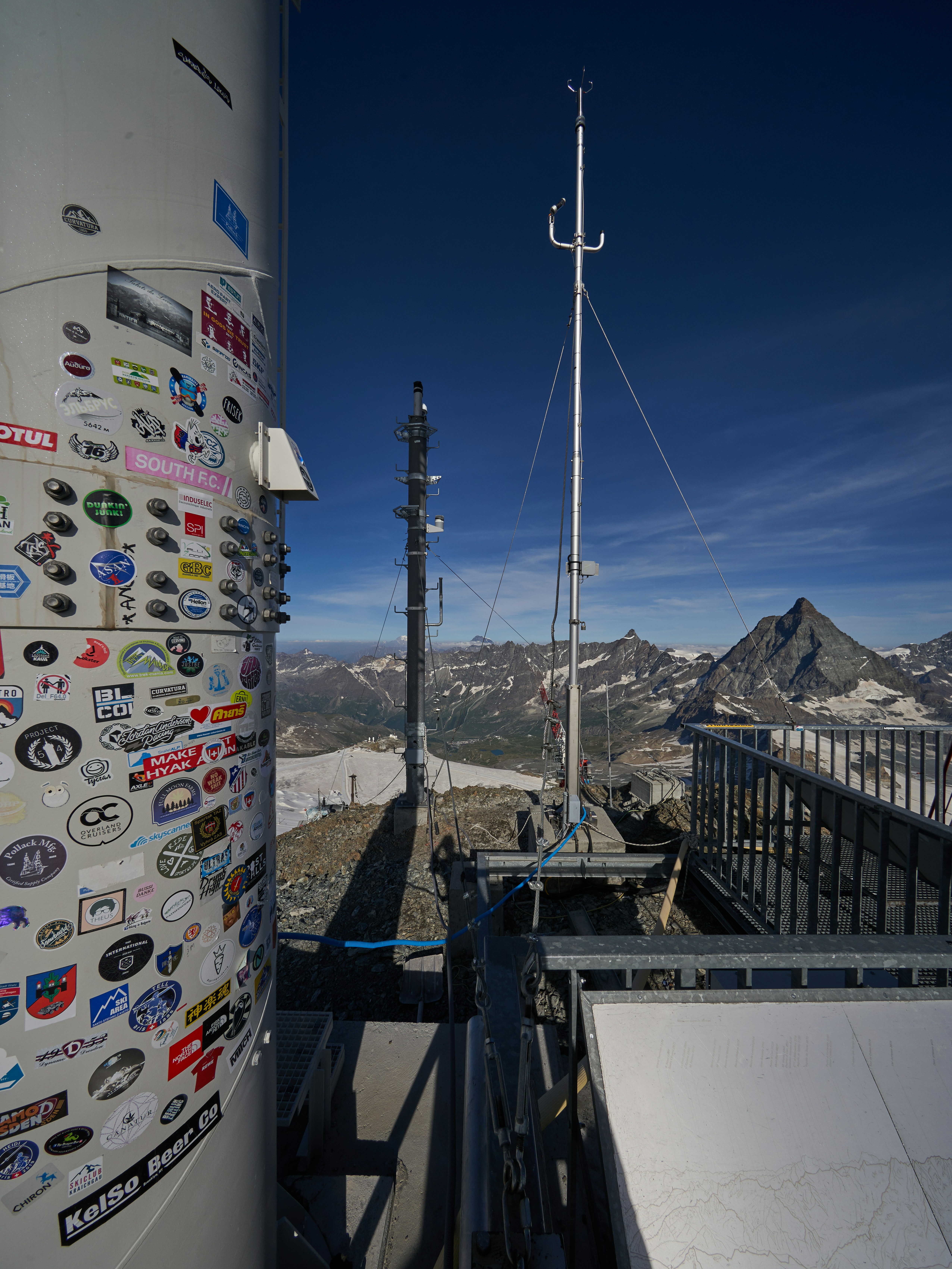A weather station adorned with various stickers stands against a backdrop of majestic mountains and a clear blue sky.