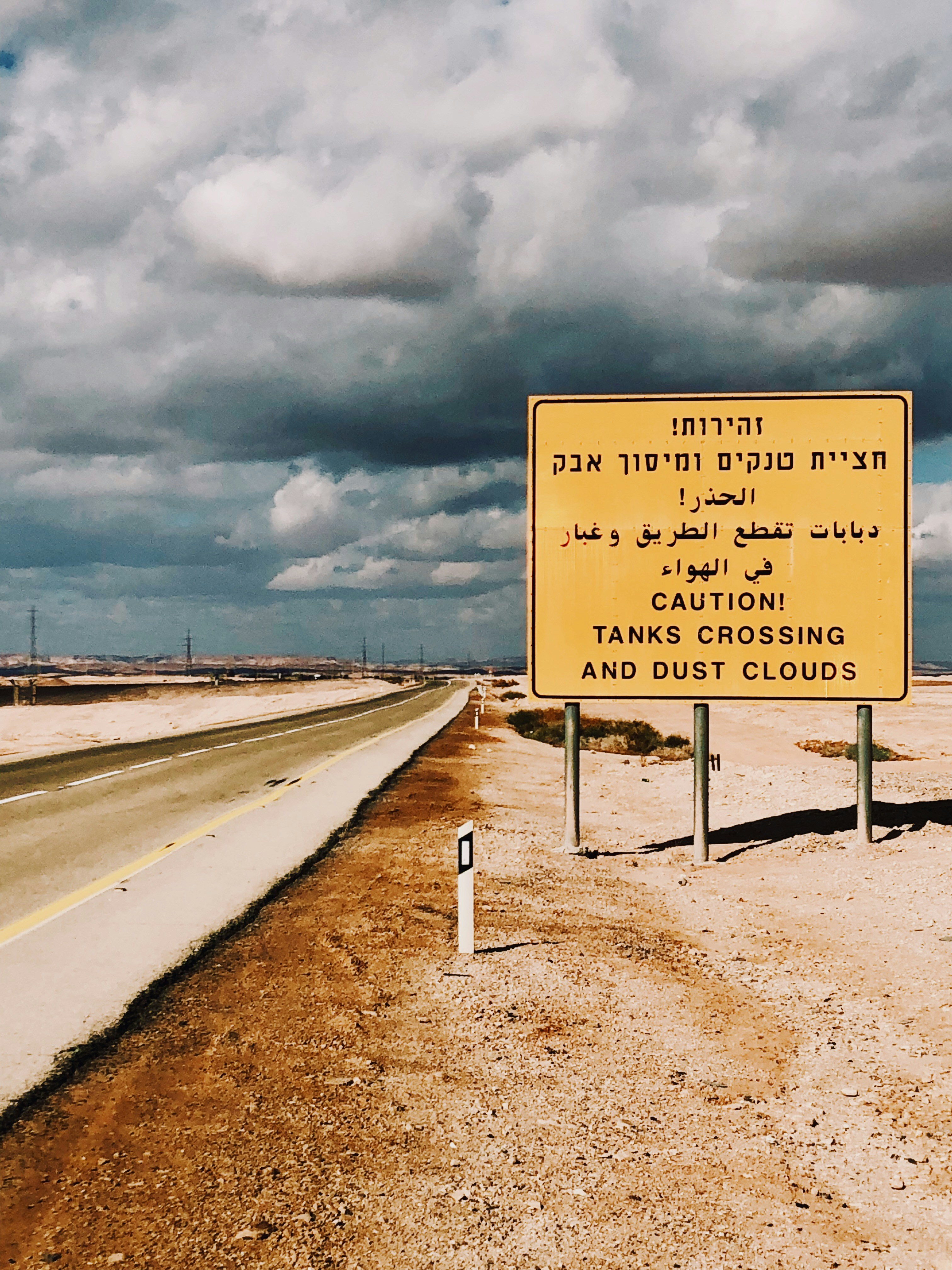 Cautionary sign in a desert landscape warning of tank crossings and dust clouds, set against a dramatic sky.