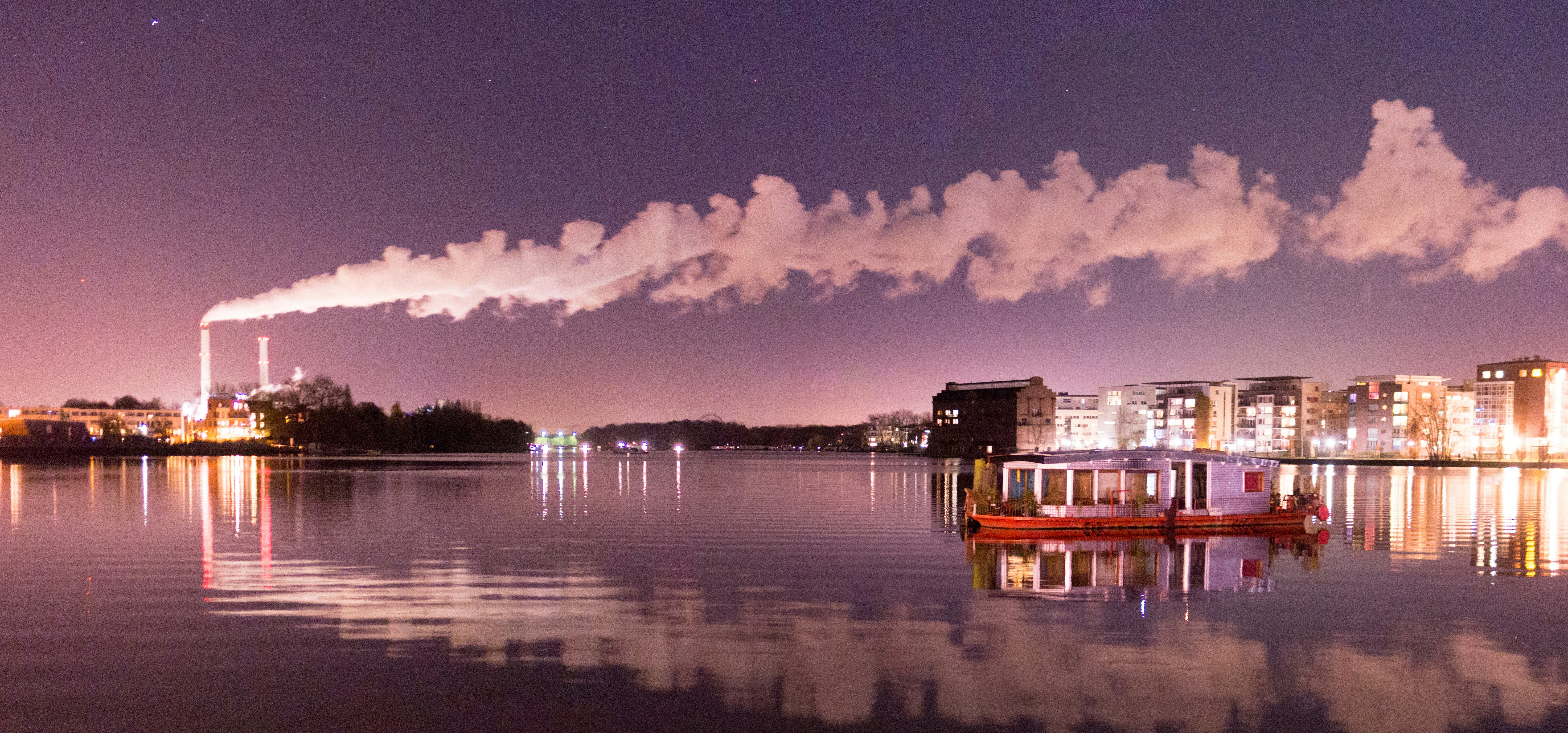 A boat floats on calm waters, reflecting the industrial skyline and smoke rising from a nearby factory against a twilight sky.
