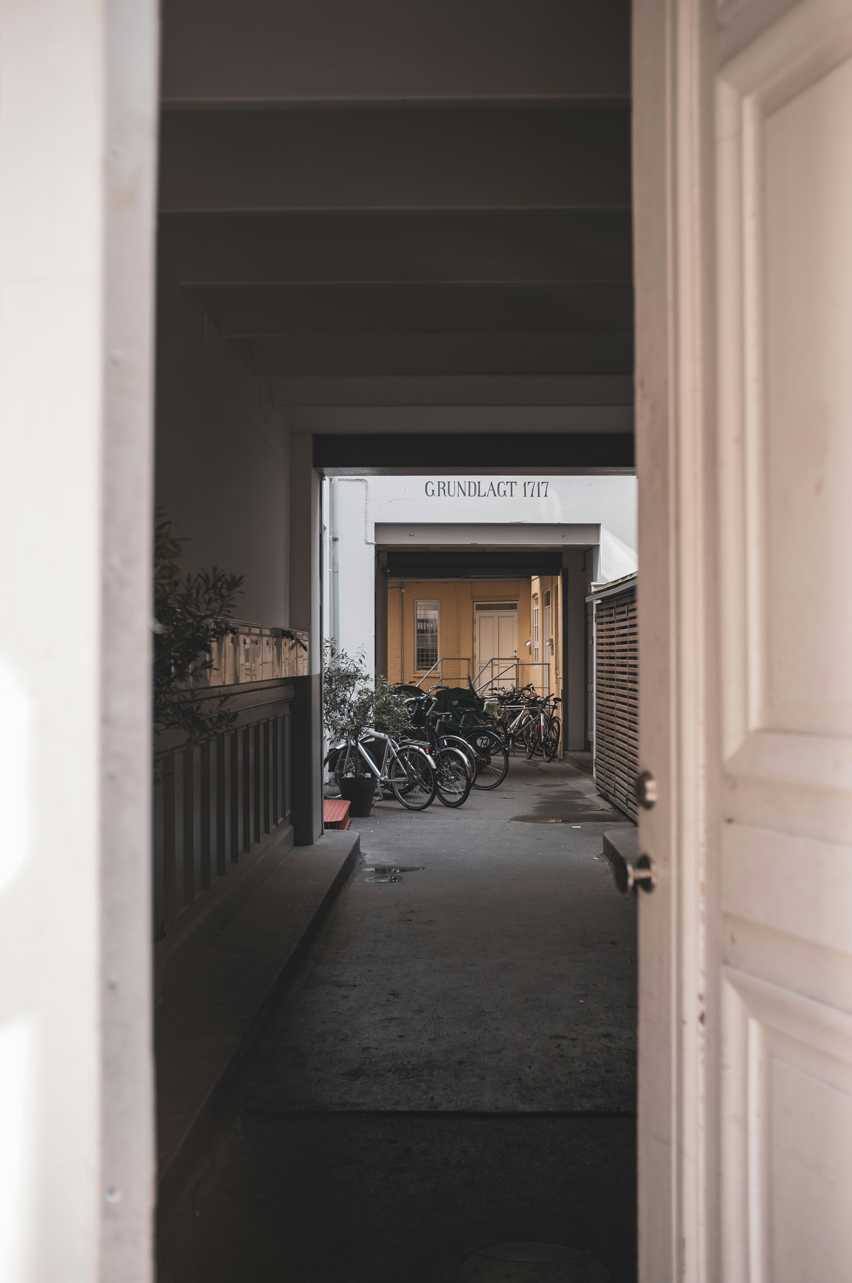 A narrow passageway leading to a yellow building, framed by an open door. Bicycles are parked neatly along the side.