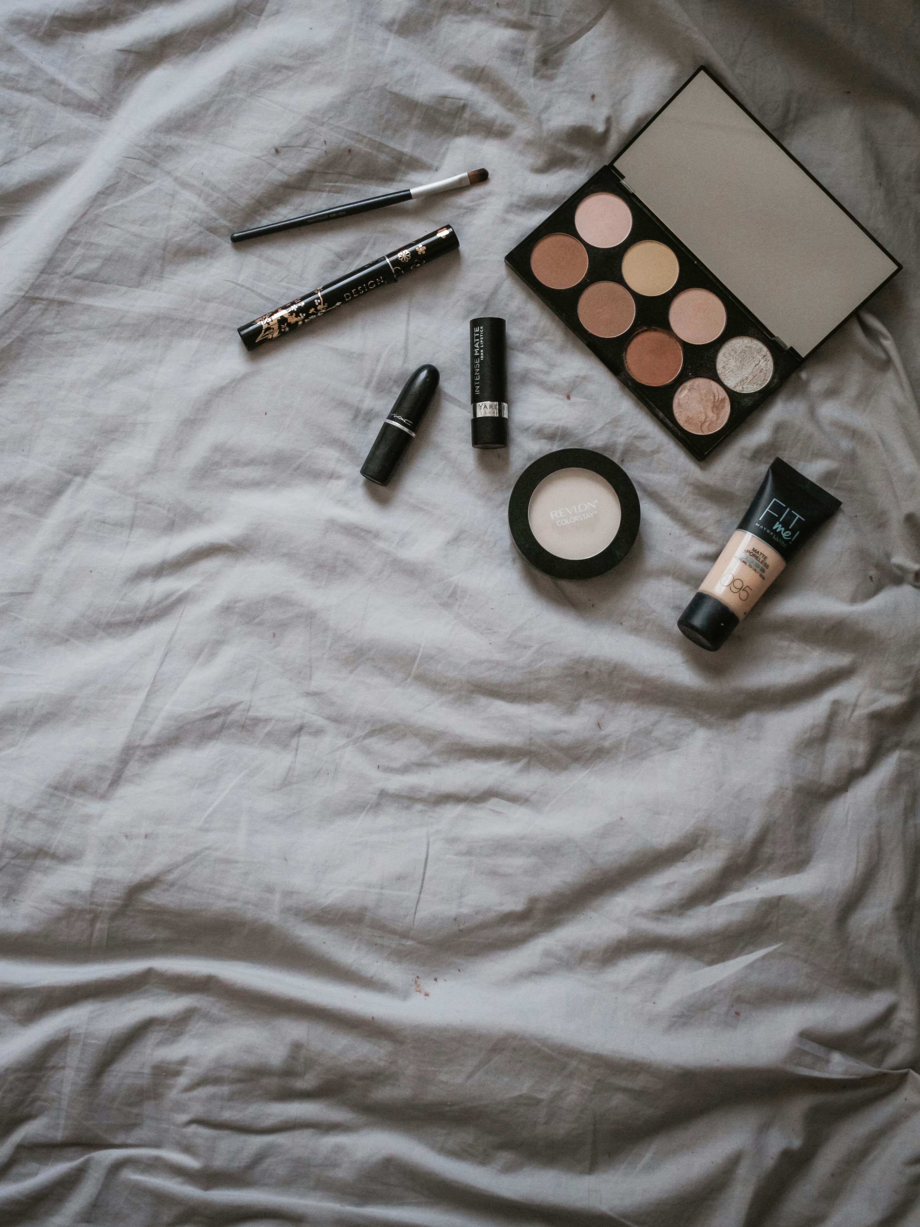 Makeup products artfully arranged on a soft, crumpled bedspread, showcasing a blend of cosmetics including a palette, lipsticks, and foundation. 