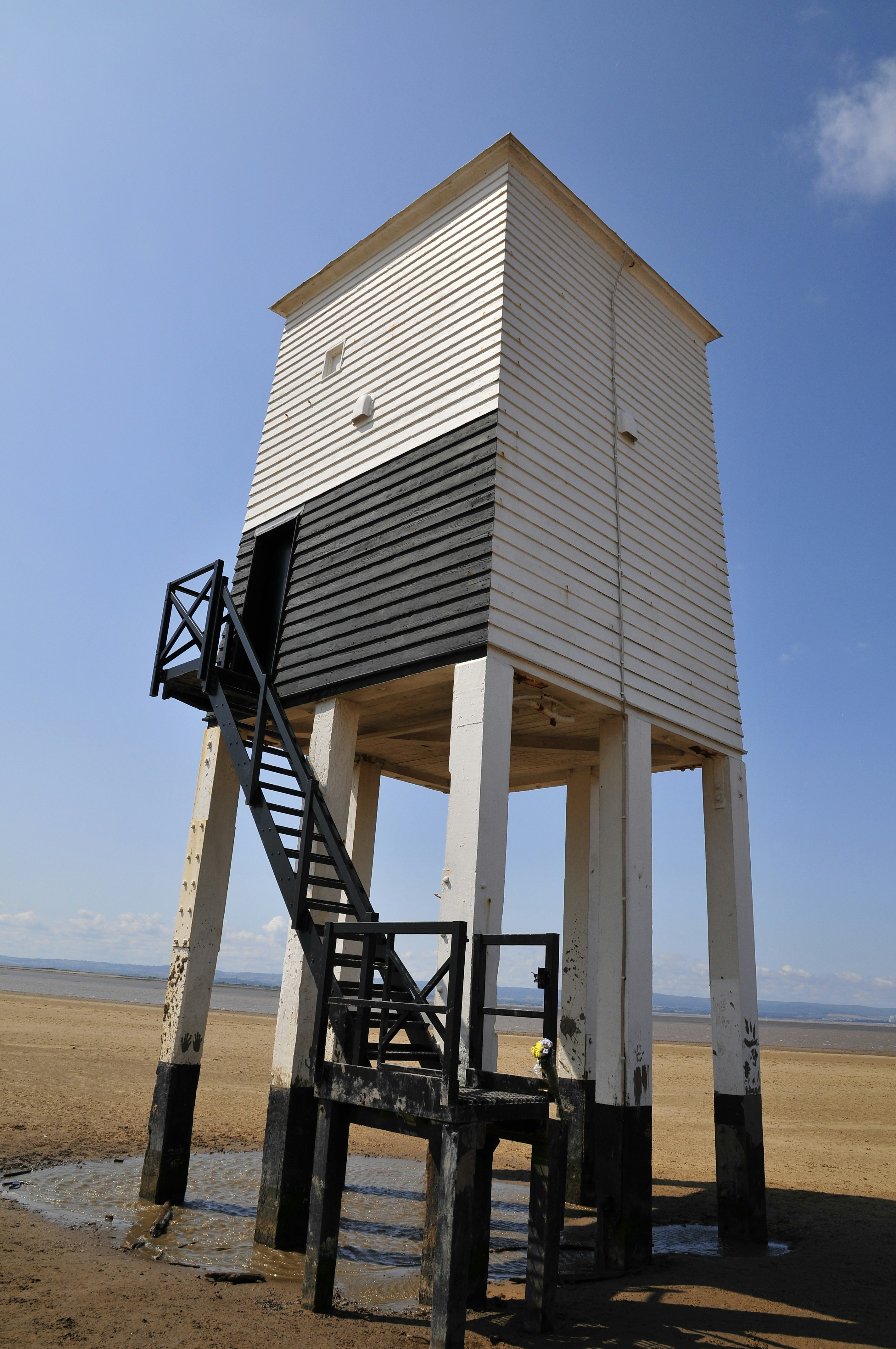 A tall, white beach watchtower perched on stilts, with a staircase leading up to the entrance, surrounded by a vast expanse of sand and a serene sky.