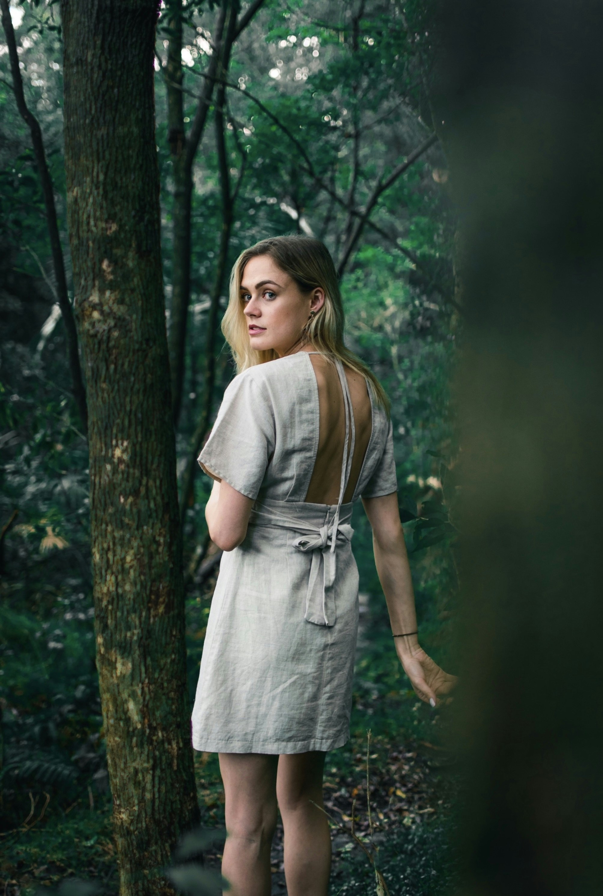 A young woman in a minimalist dress stands amidst lush greenery, capturing a moment of tranquility in nature. Her gaze over the shoulder invites curiosity.
