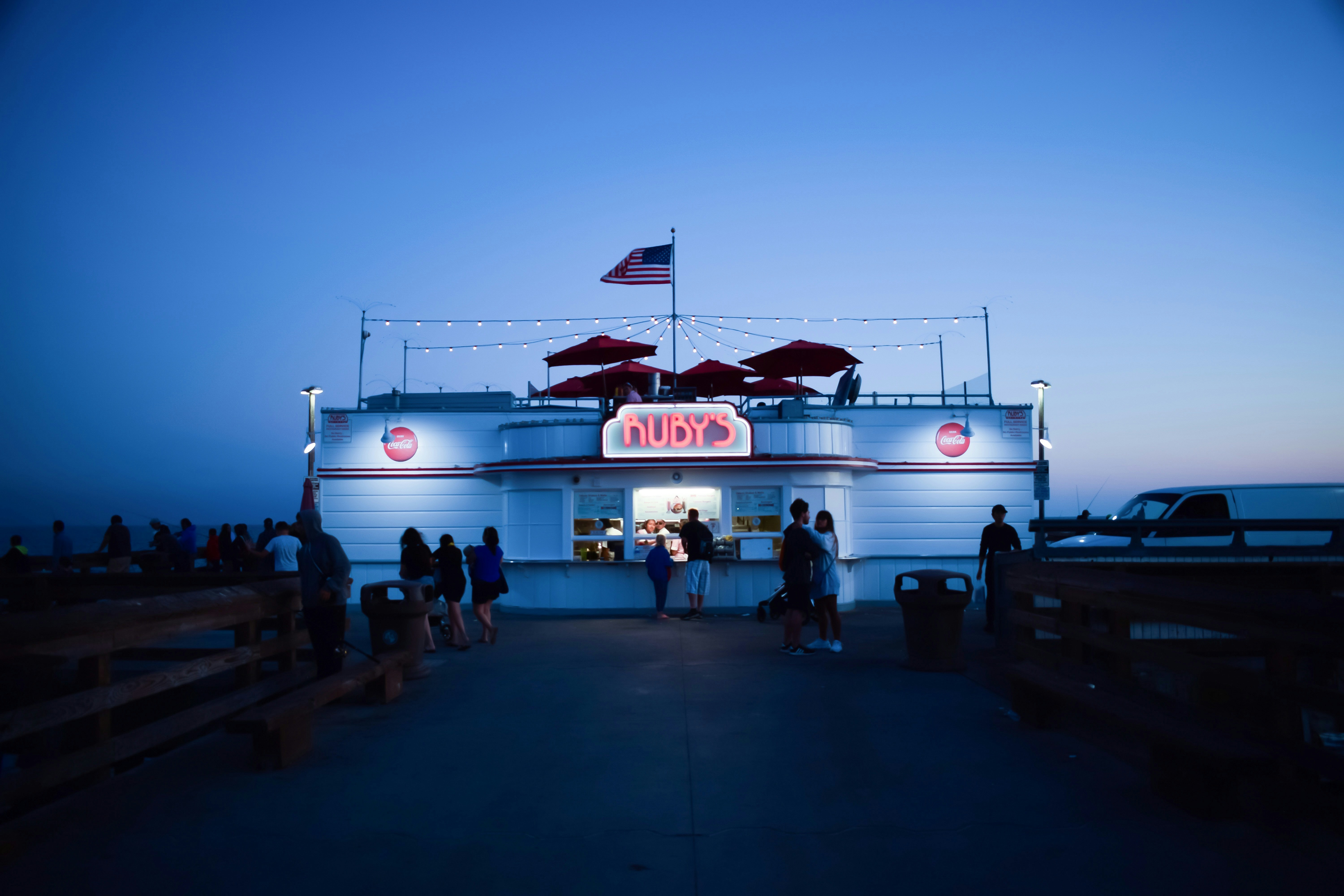 Seaside snack bar illuminated under a deep blue evening sky, with people silhouetted against the horizon.