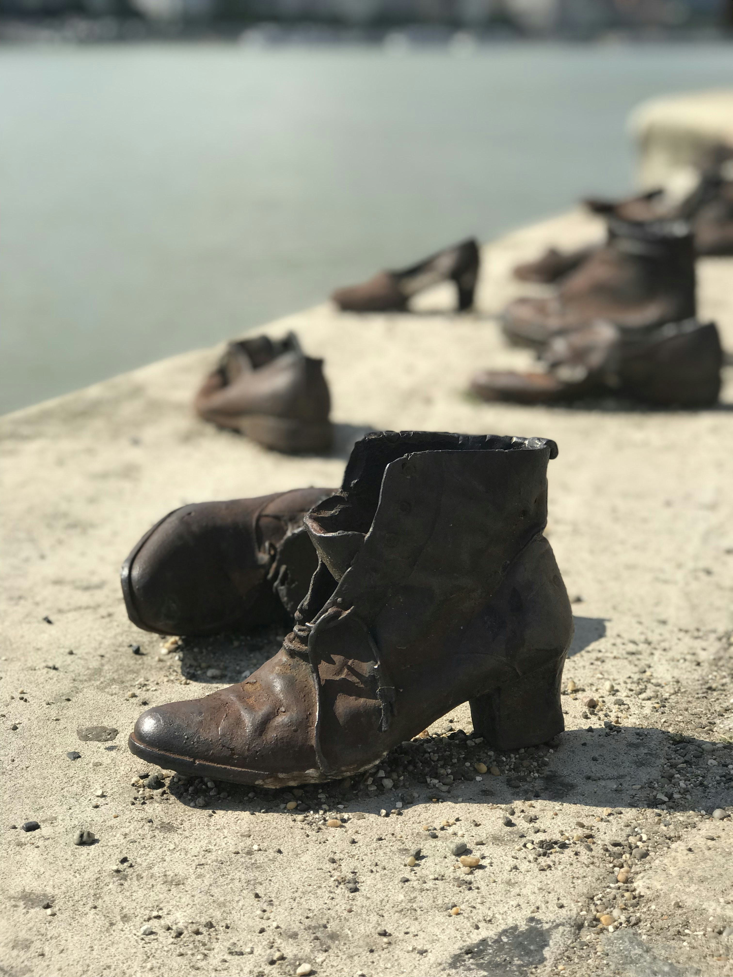 Weathered brown boots scattered along a riverbank, symbolizing loss and memory. The serene water serves as a poignant backdrop.