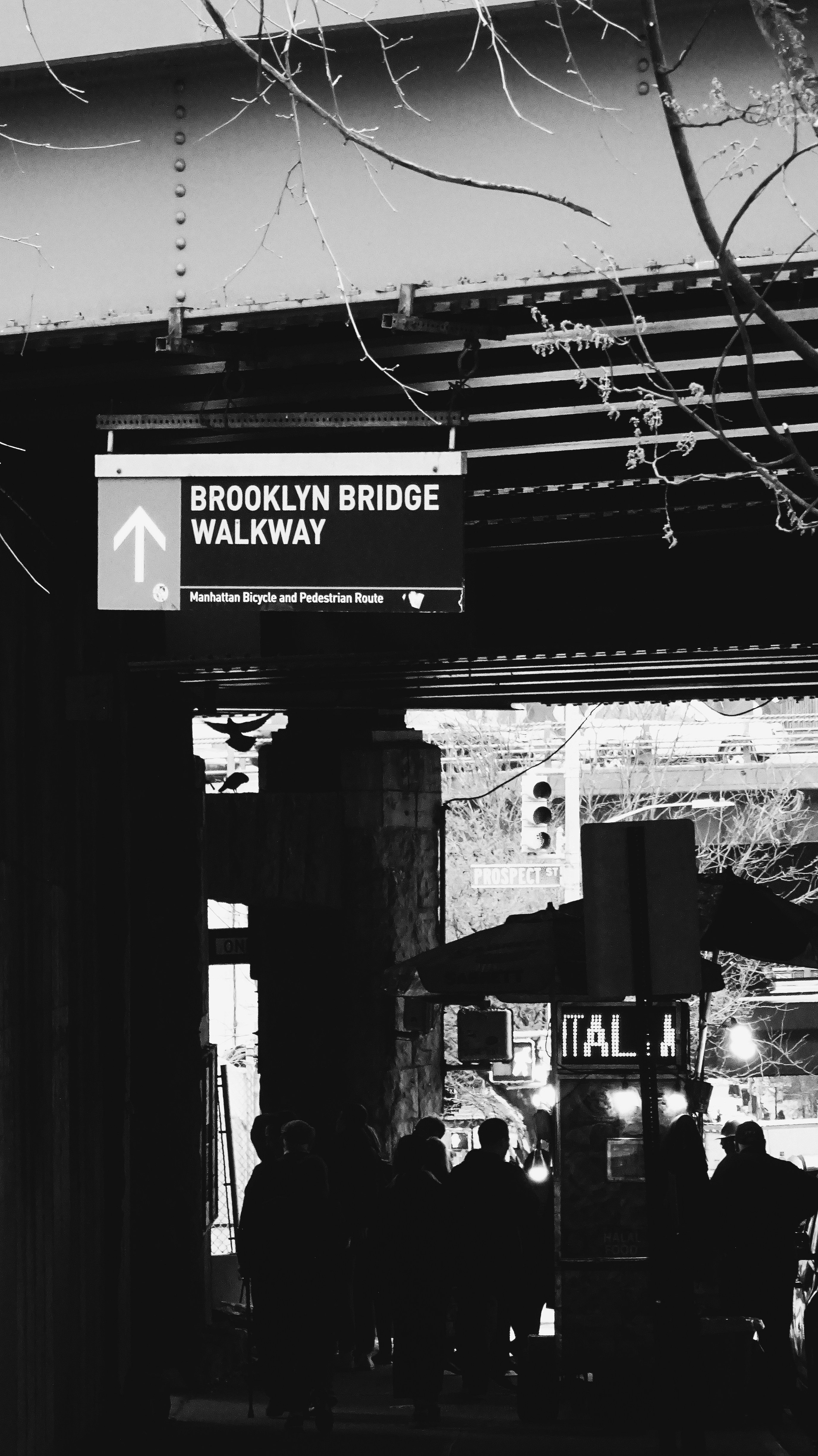 Sign directing pedestrians to the Brooklyn Bridge walkway under an urban overpass, with silhouettes of people in the foreground. 