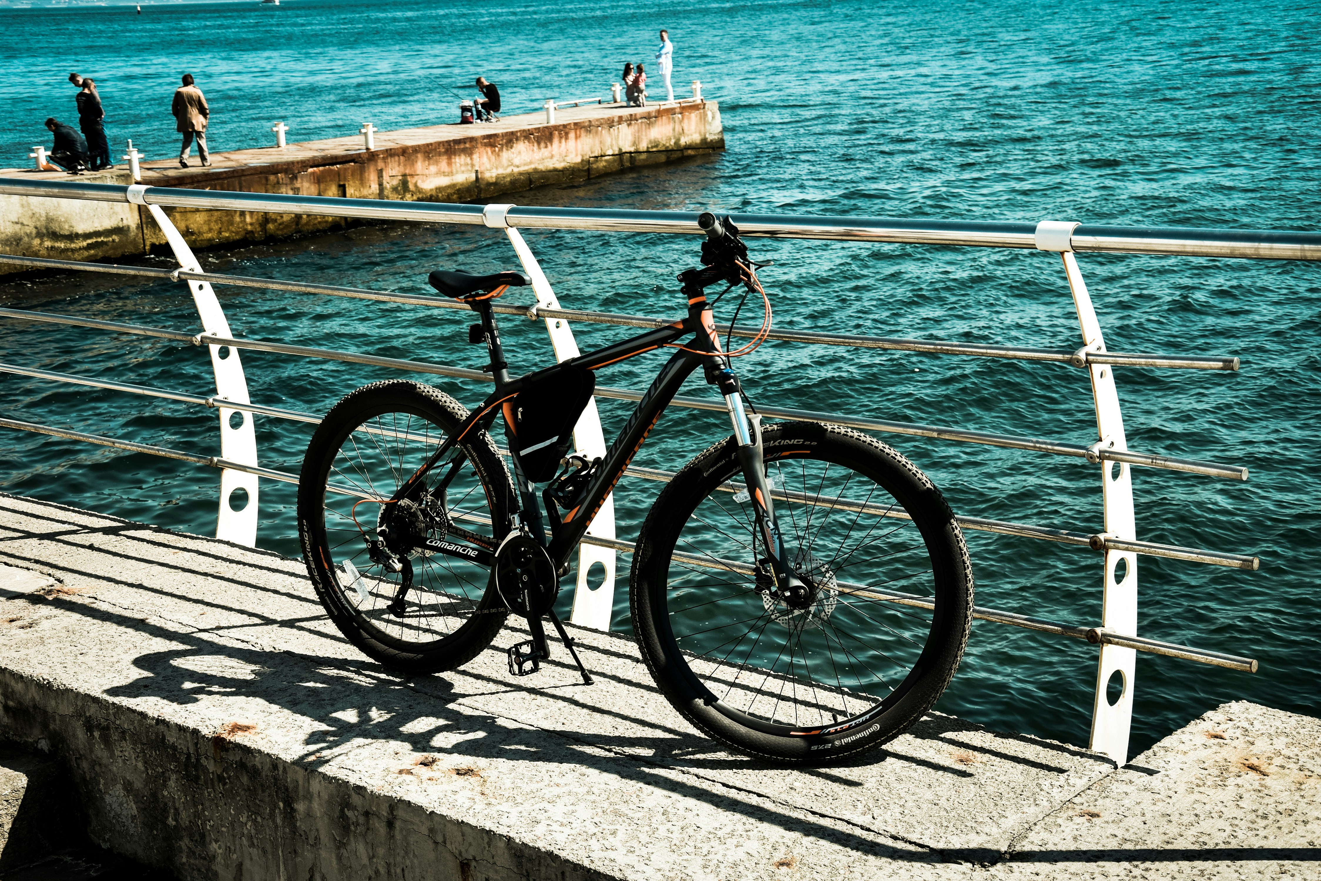 Mountain bike resting against a modern railing near a vibrant waterfront, with people enjoying the view in the background.