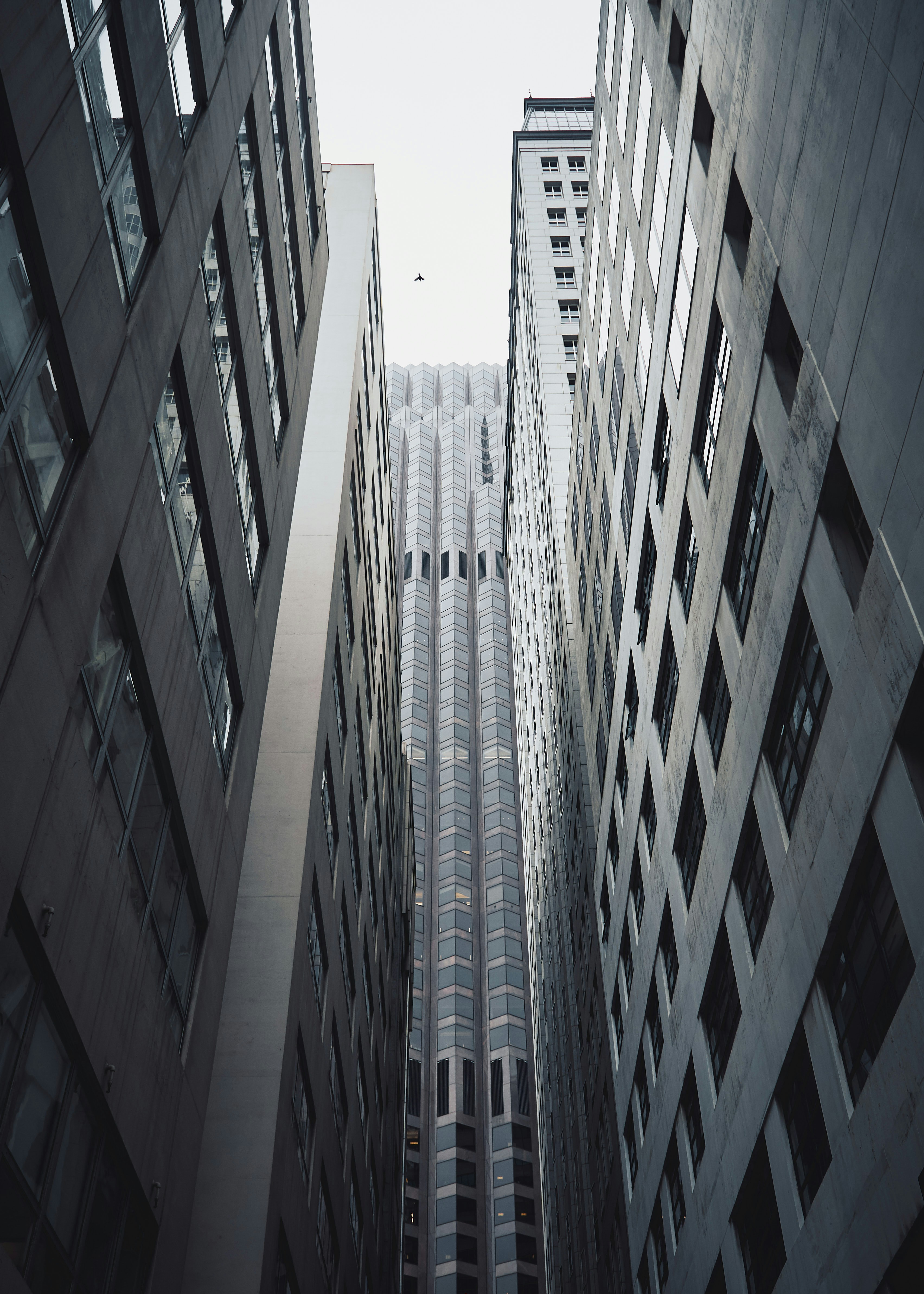 View looking up between towering skyscrapers, revealing a glimpse of a distant building against a gray sky.