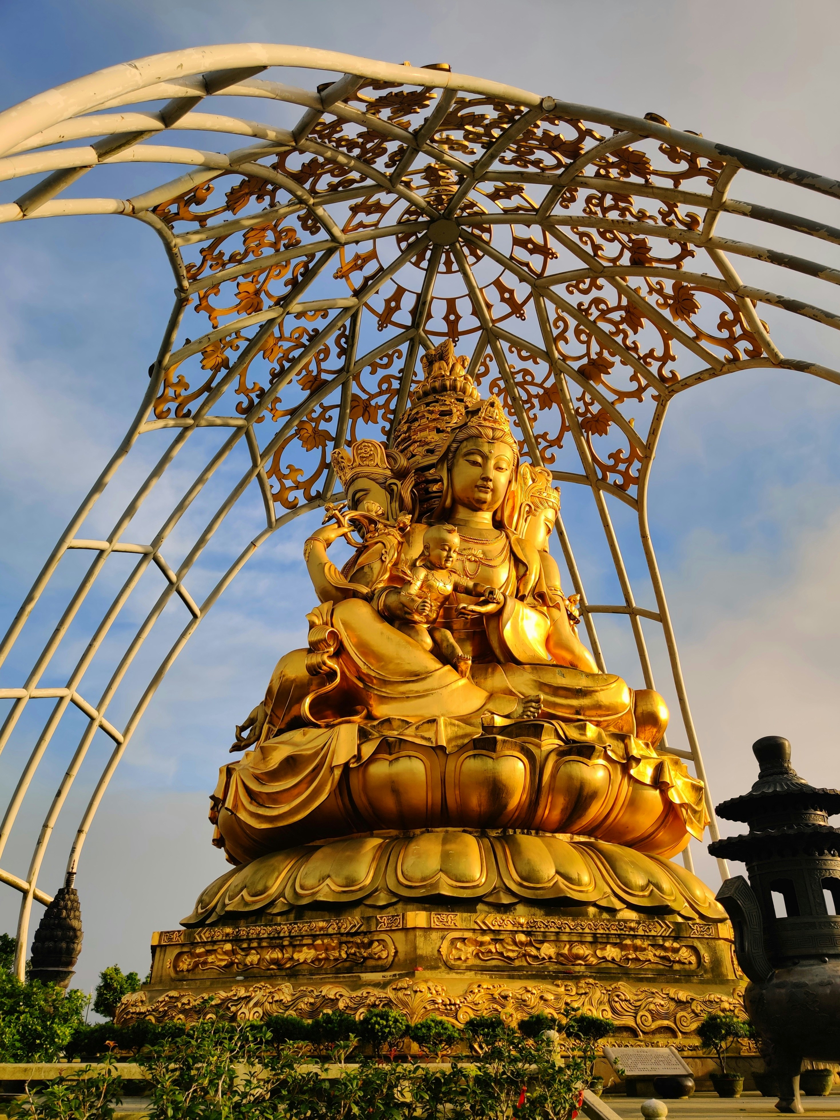 A gilded Buddhist statue seated in lotus pose beneath an ornate lattice canopy, set against a clear blue sky.