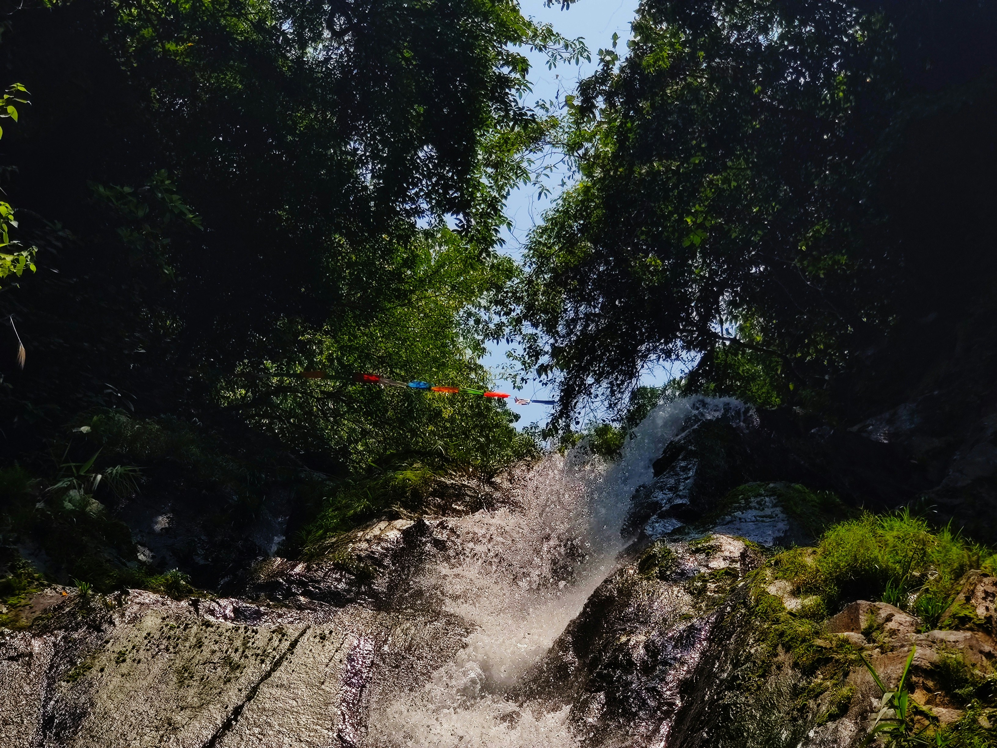 Waterfall tumbling down rocky terrain, surrounded by lush greenery and colorful prayer flags fluttering in the breeze.