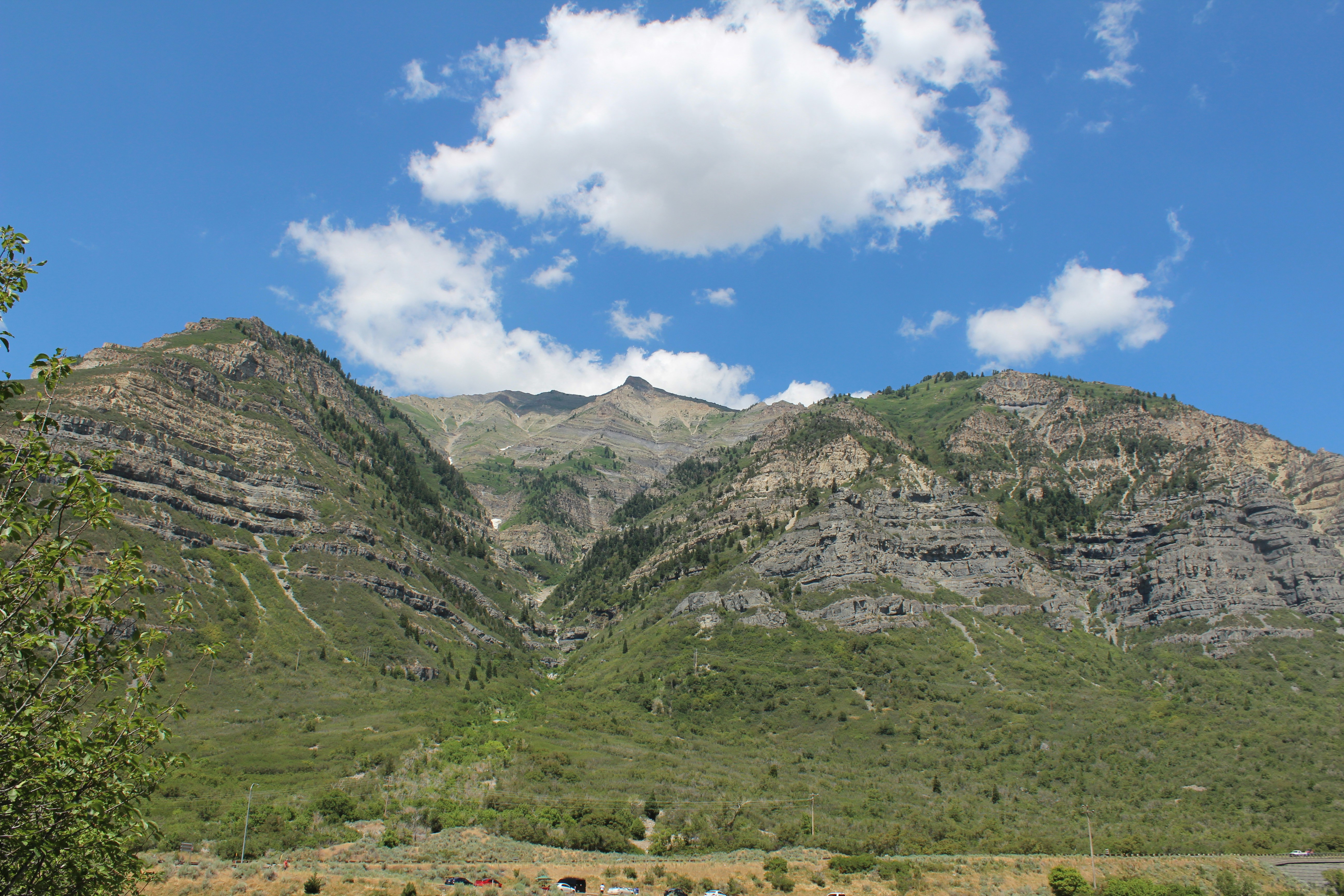 Majestic mountain range under a bright blue sky with fluffy clouds, showcasing layered rock formations and lush greenery at the base.