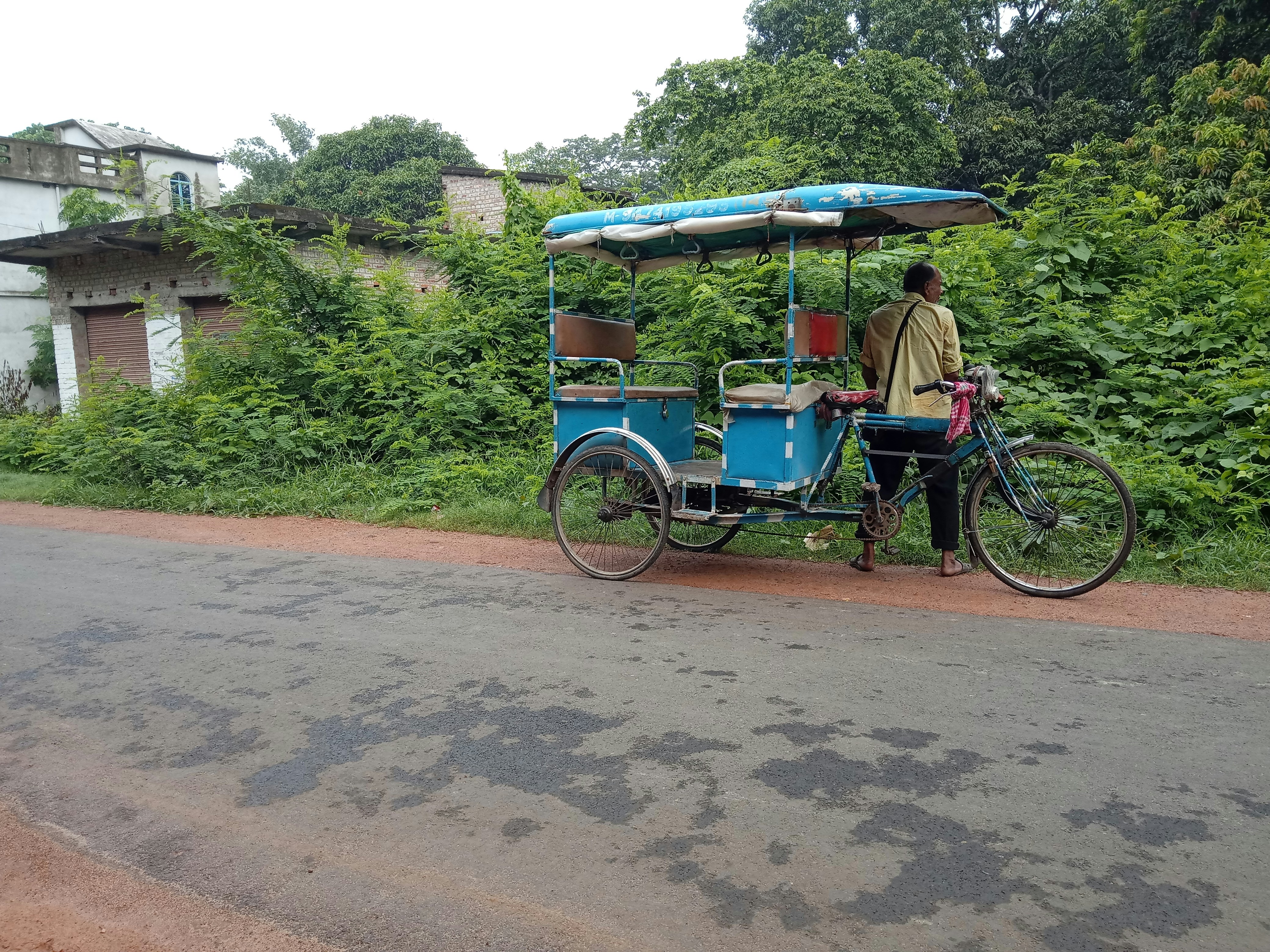 Street vendor beside a blue pedal cart on a paved road, with lush greenery in the background.