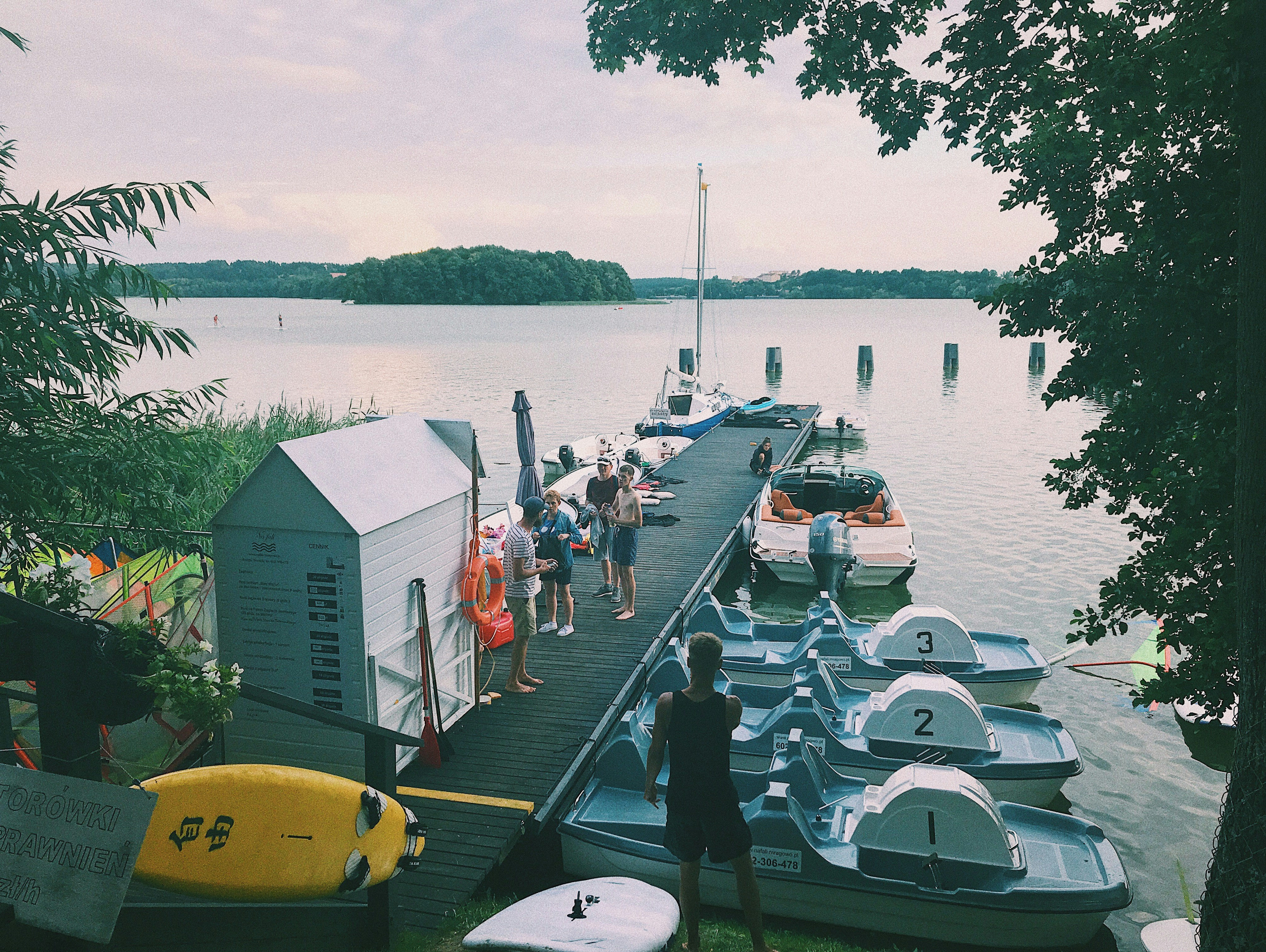People gather on a wooden dock lined with paddle boats, set against a tranquil lake and overcast sky.