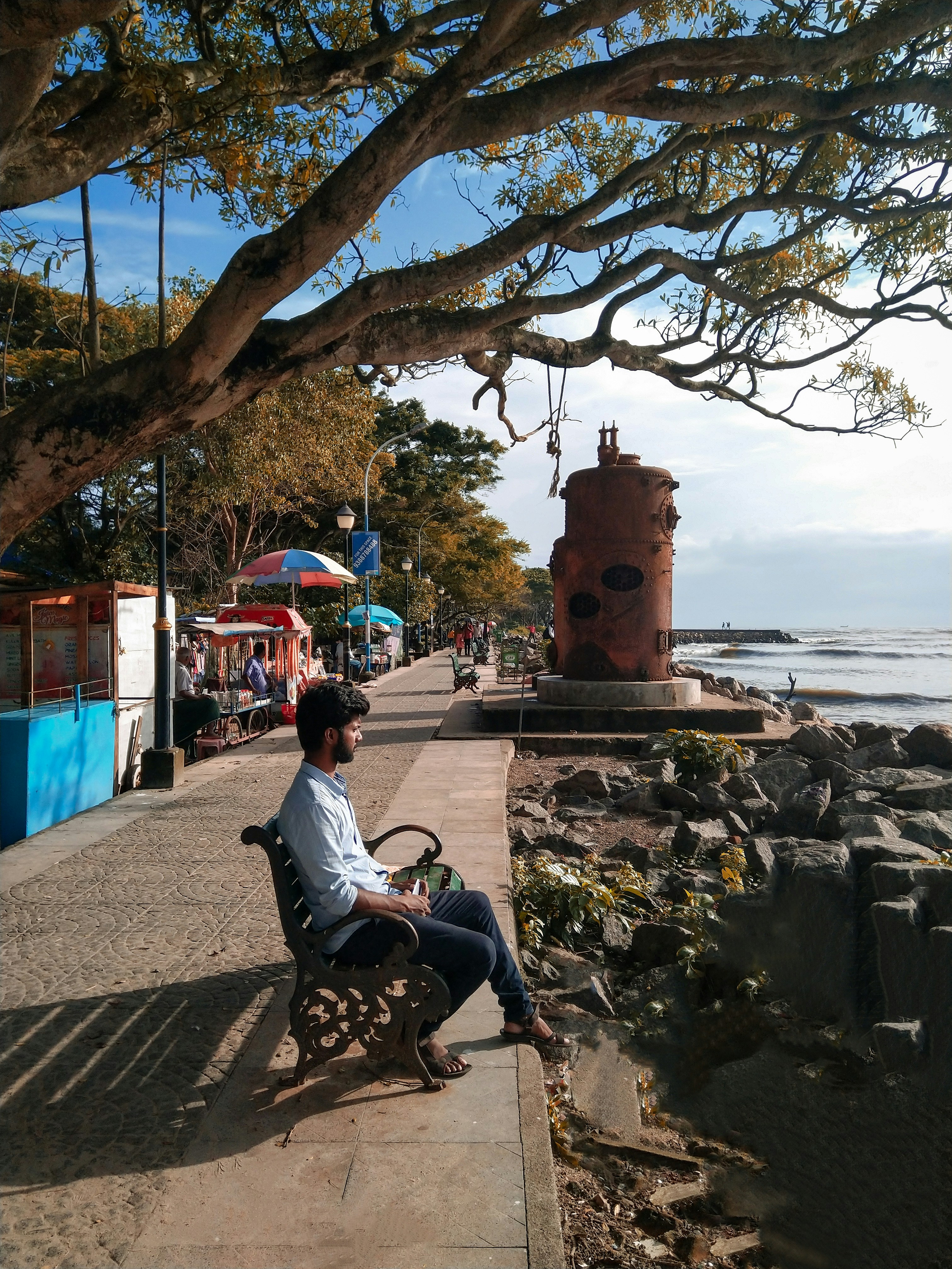 A man seated on a bench along a coastal promenade, with an old submarine structure nearby and vibrant umbrellas in the background. The scene captures a serene moment by the water.