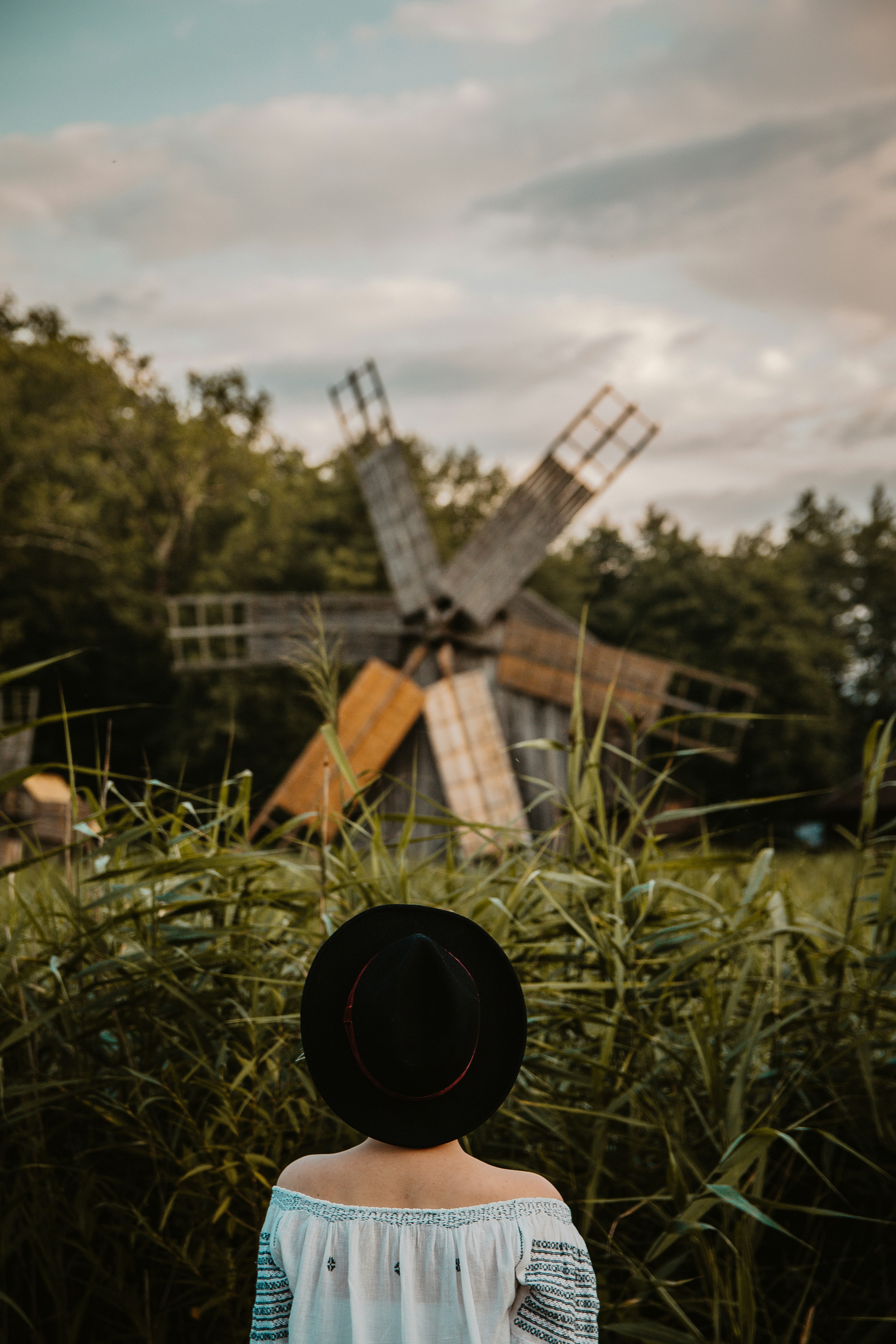 Person gazes at an old windmill surrounded by tall grass under a cloudy sky, evoking nostalgia and connection to history.