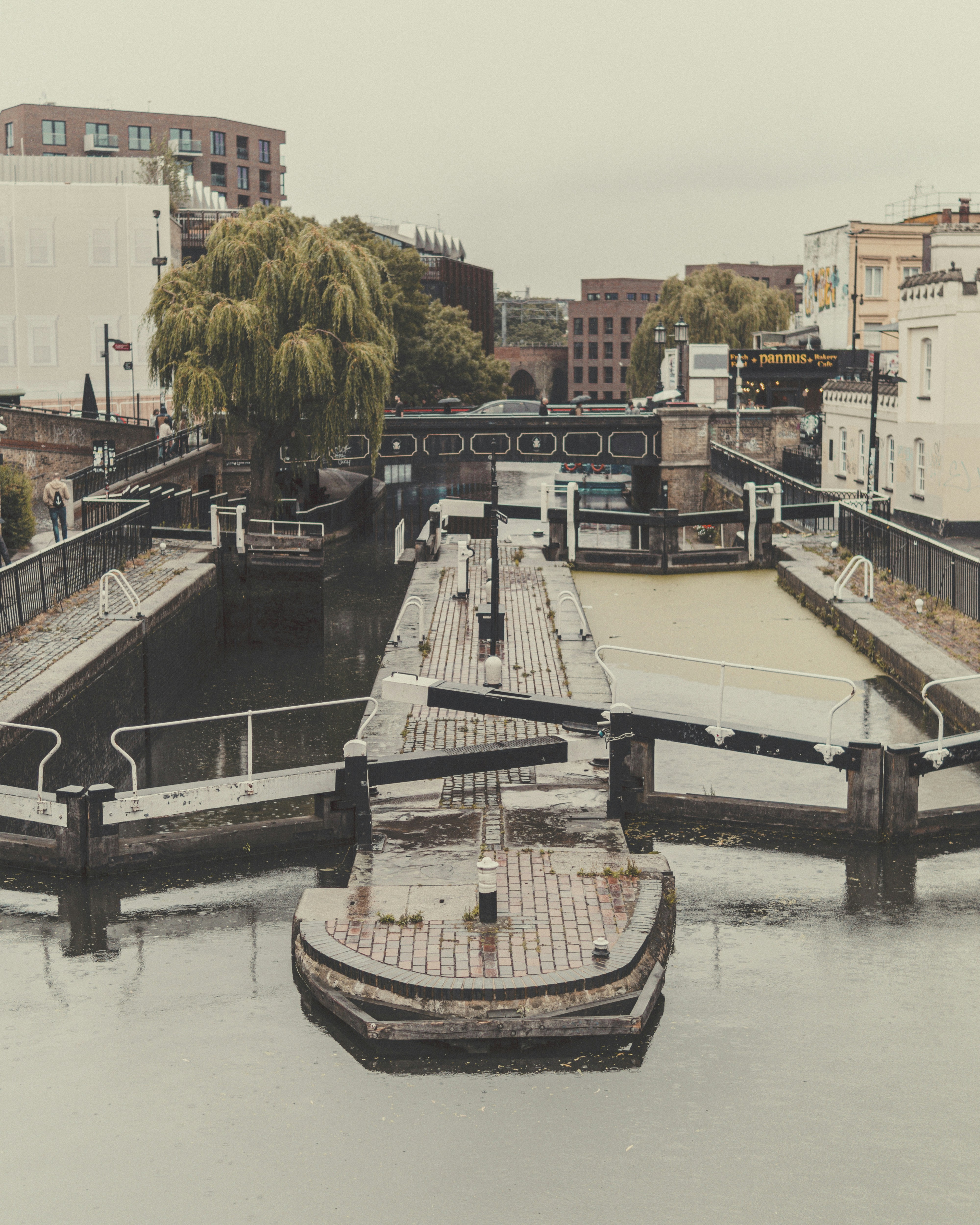 Quaint canal lock surrounded by historic buildings and lush trees under an overcast sky.