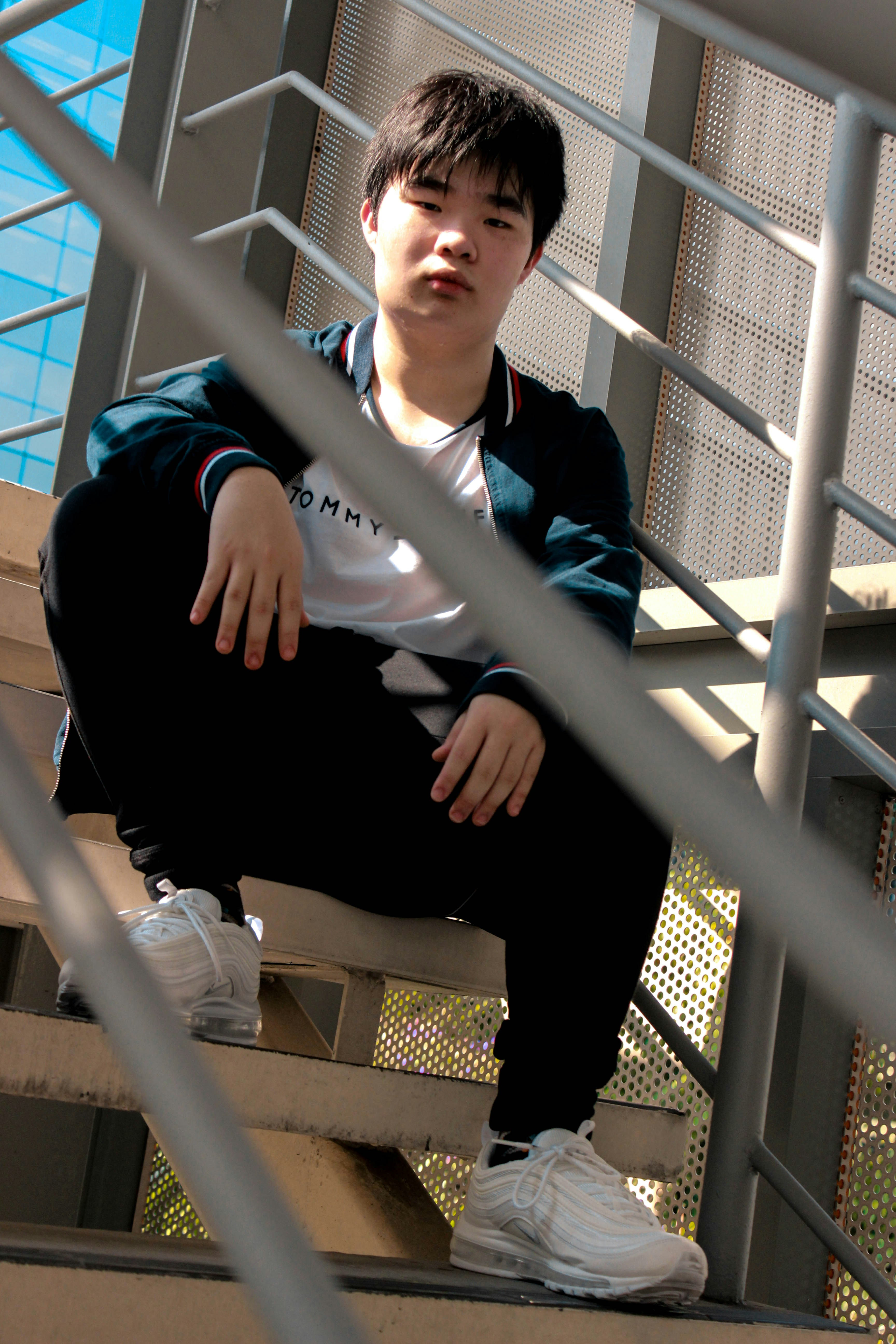 Young person sitting on stairs in an urban setting, framed by metallic railings. The scene captures a blend of modern architecture and casual style.