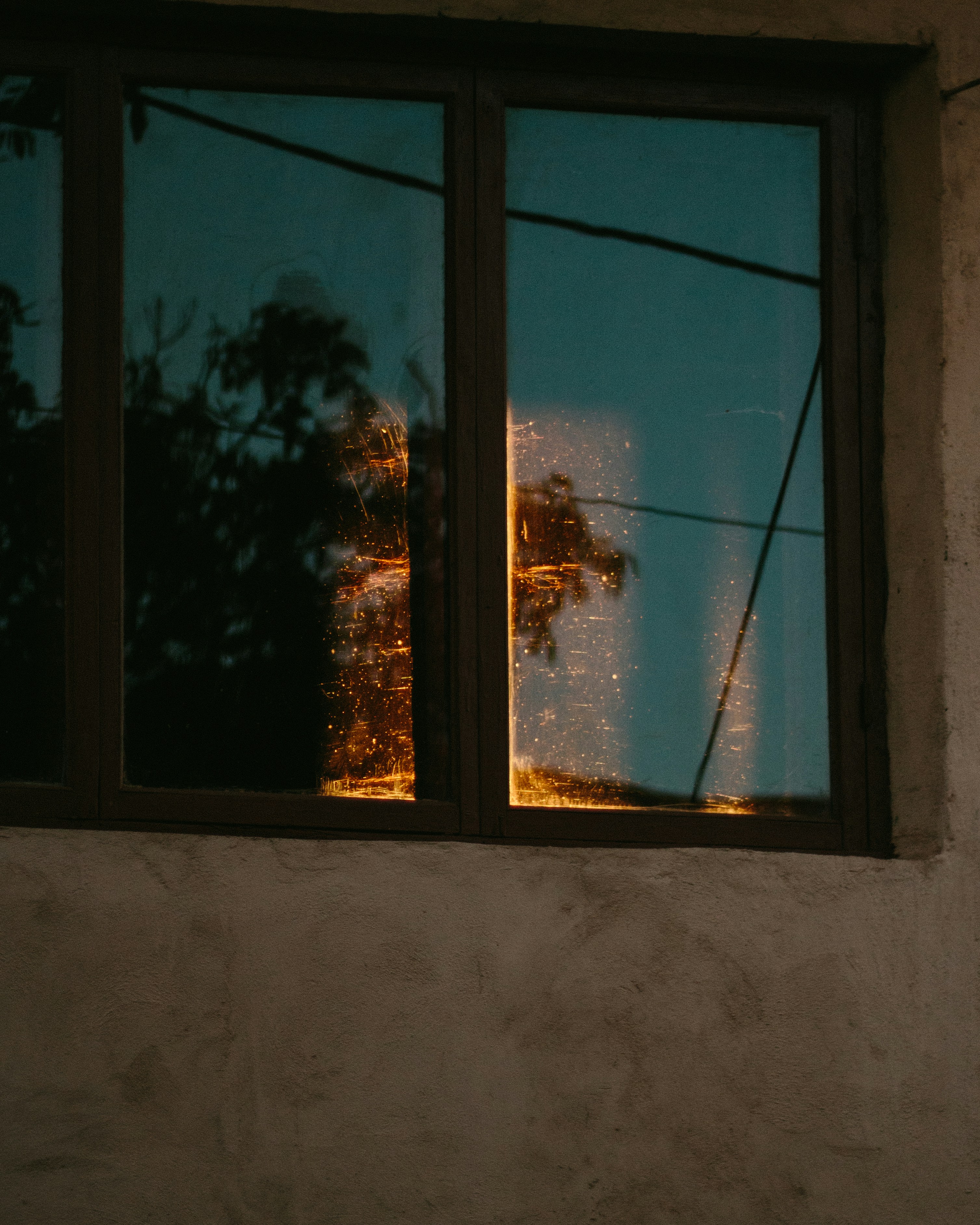 A close-up view of a window reflecting the warm glow of lights against a darkening sky, framed by the textured wall of a building.