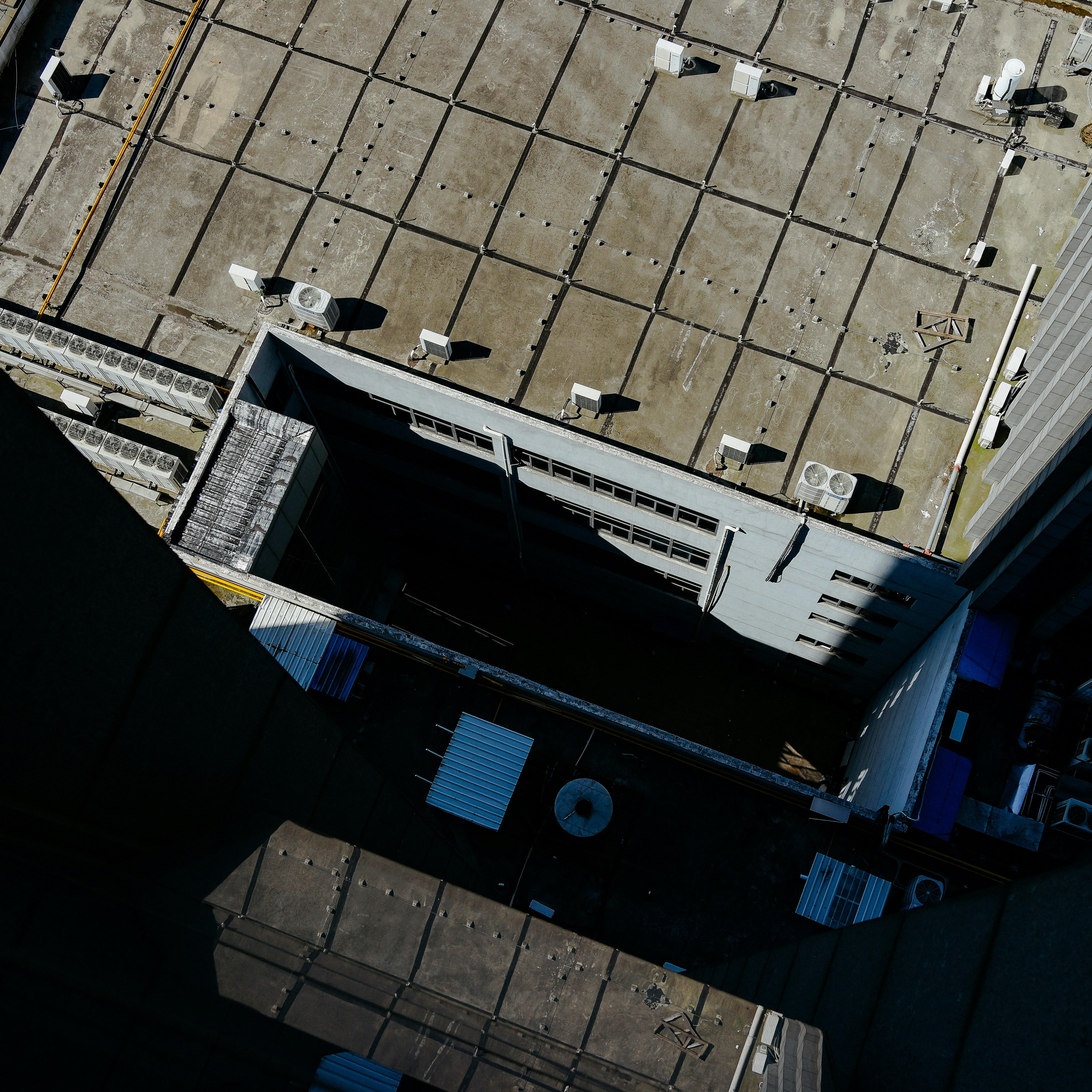 High-angle photograph of a flat rooftop with a geometric grid pattern, scattered HVAC units, and strong shadows between buildings.