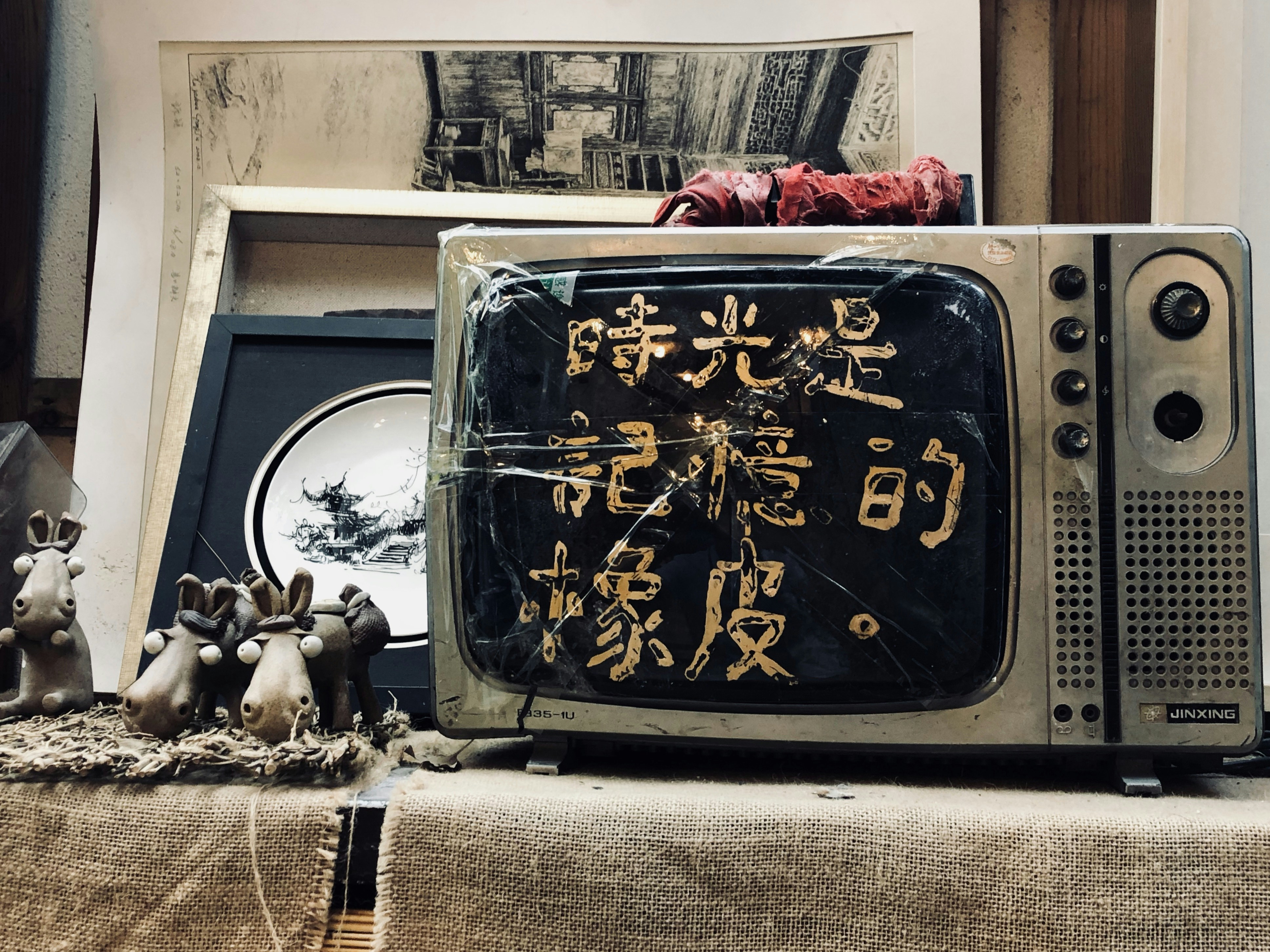 An aged television set adorned with Chinese characters, surrounded by rustic decor including a framed artwork and small figurines. The scene evokes a sense of nostalgia.