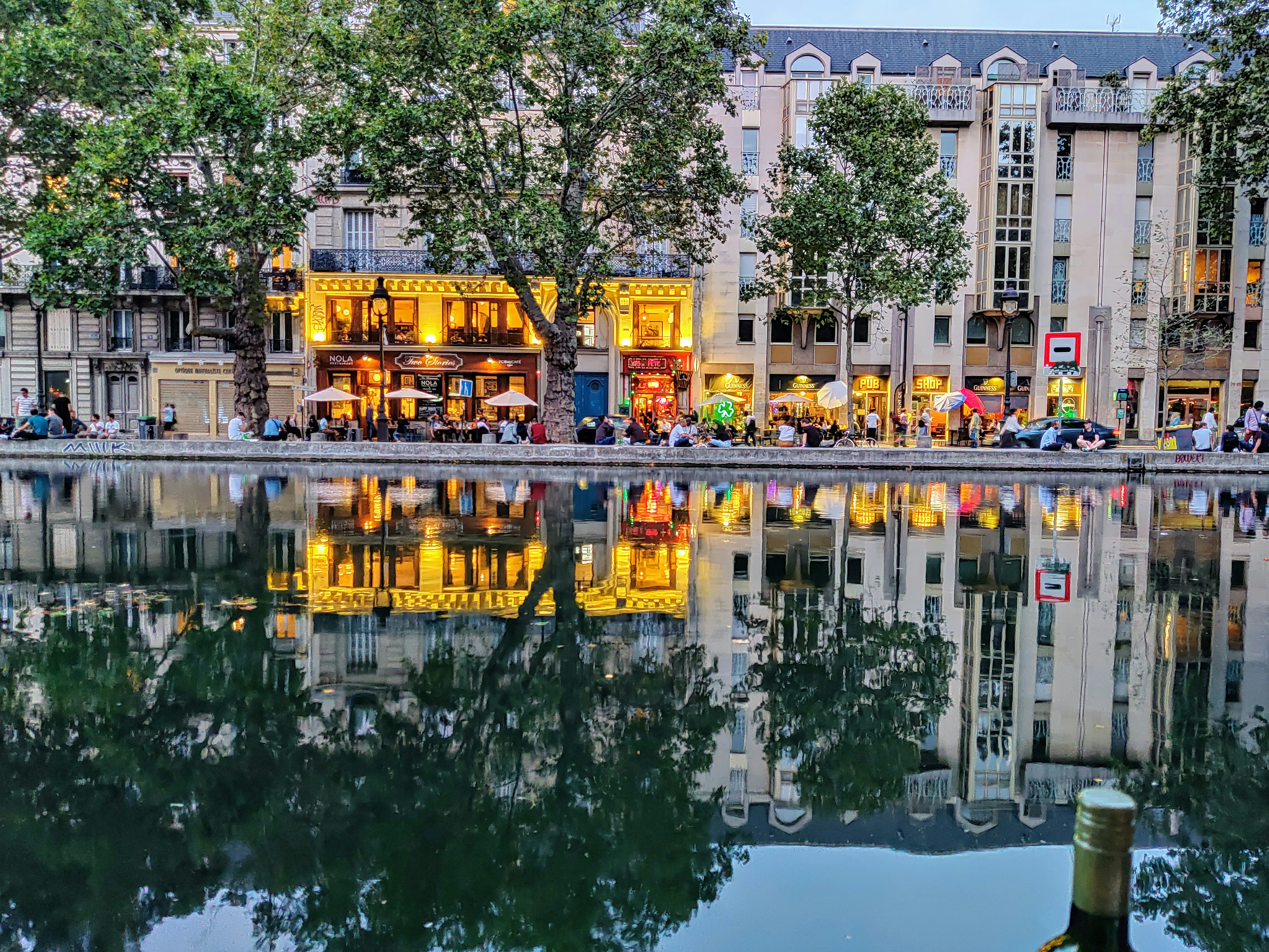 Charming waterfront scene featuring restaurants and cafes, with vibrant reflections mirrored in the calm water. The atmosphere is lively with people enjoying their evening.