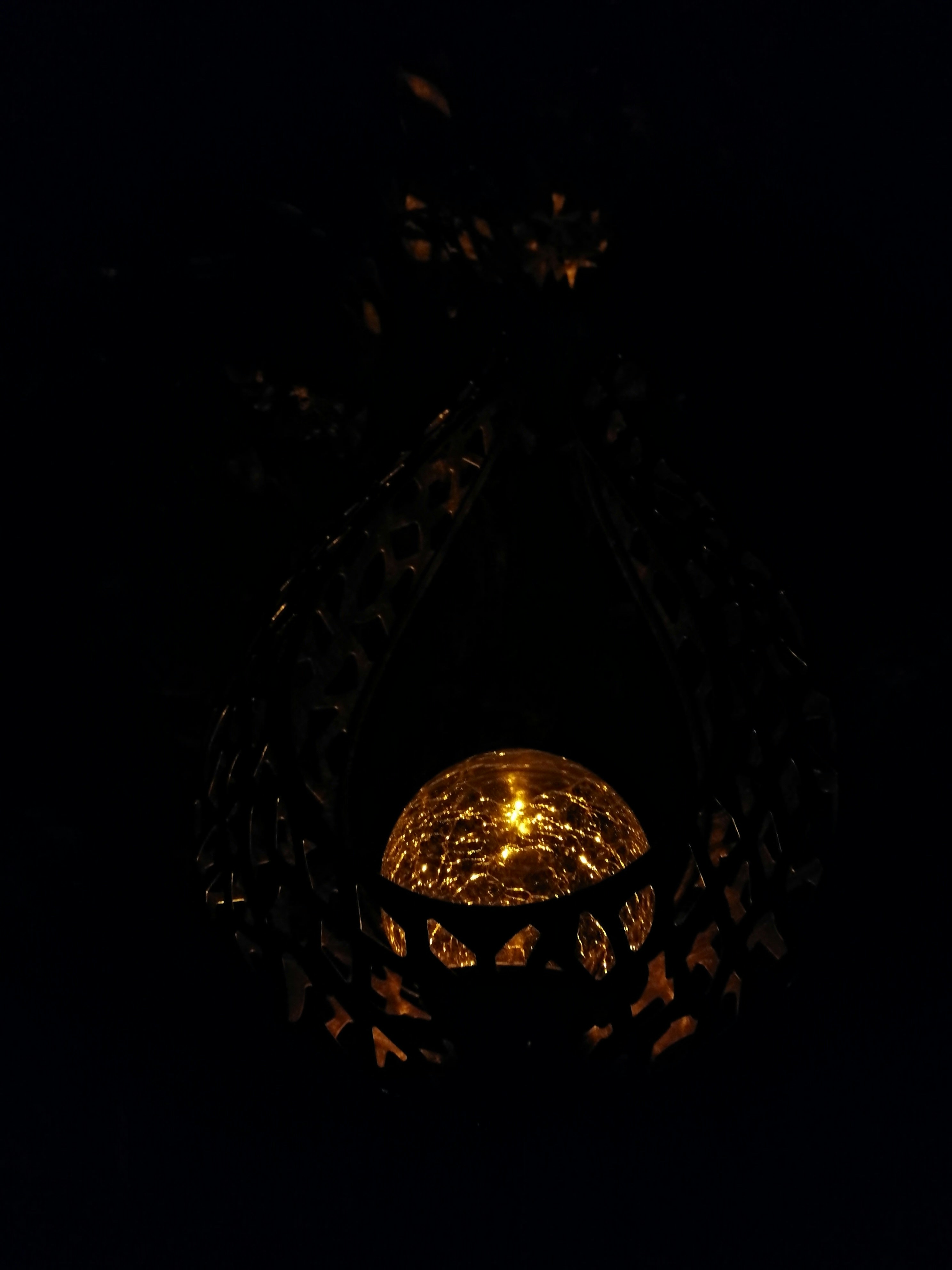A dark scene featuring a warm amber bulb enclosed by a faceted metal lantern, its lattice reflections creating intricate highlights. The glow centers attention on the orb while the surroundings remain largely in shadow.